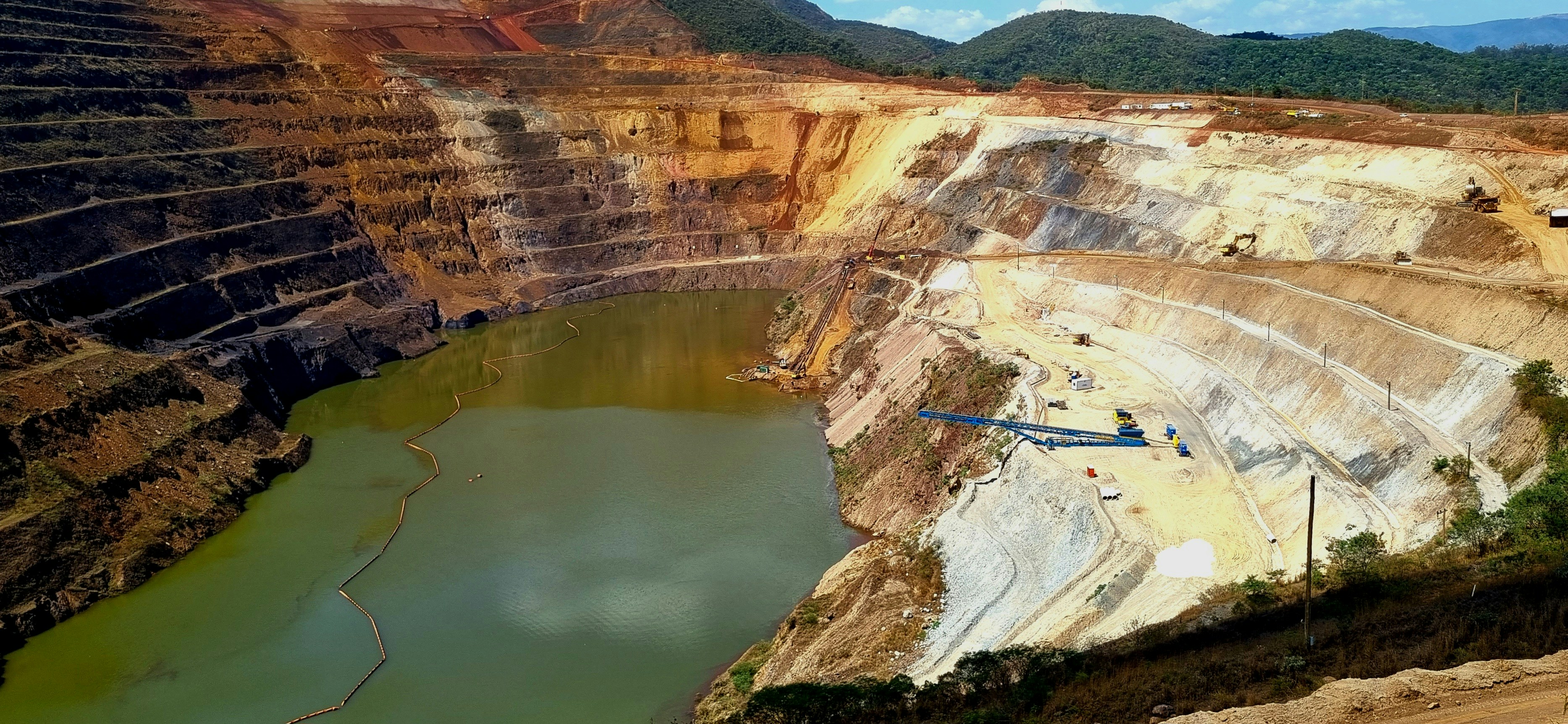 Expansive open-pit mine with terraced layers and a green water basin, surrounded by rugged hills.