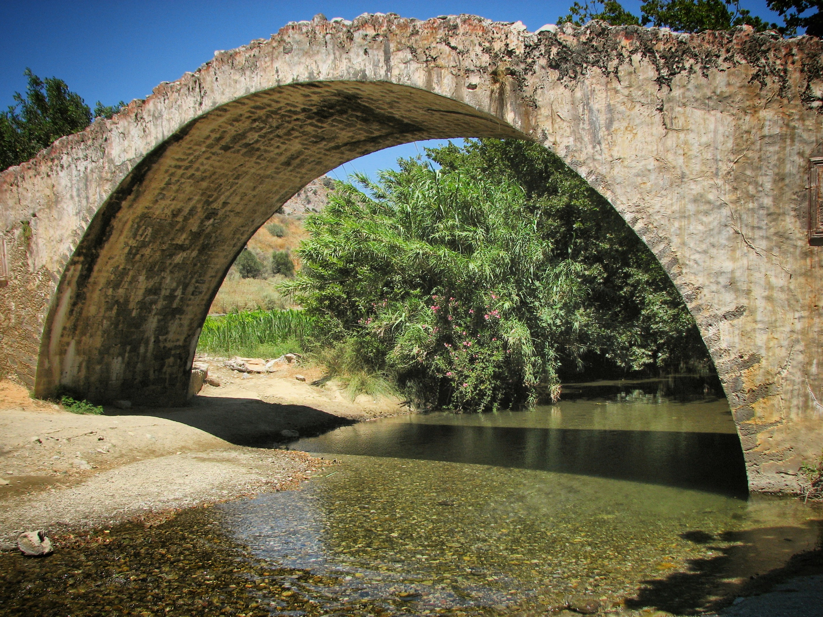 Weathered stone arch spans a calm river, framed by lush greenery. A daylight photograph capturing the tranquil riverscape.