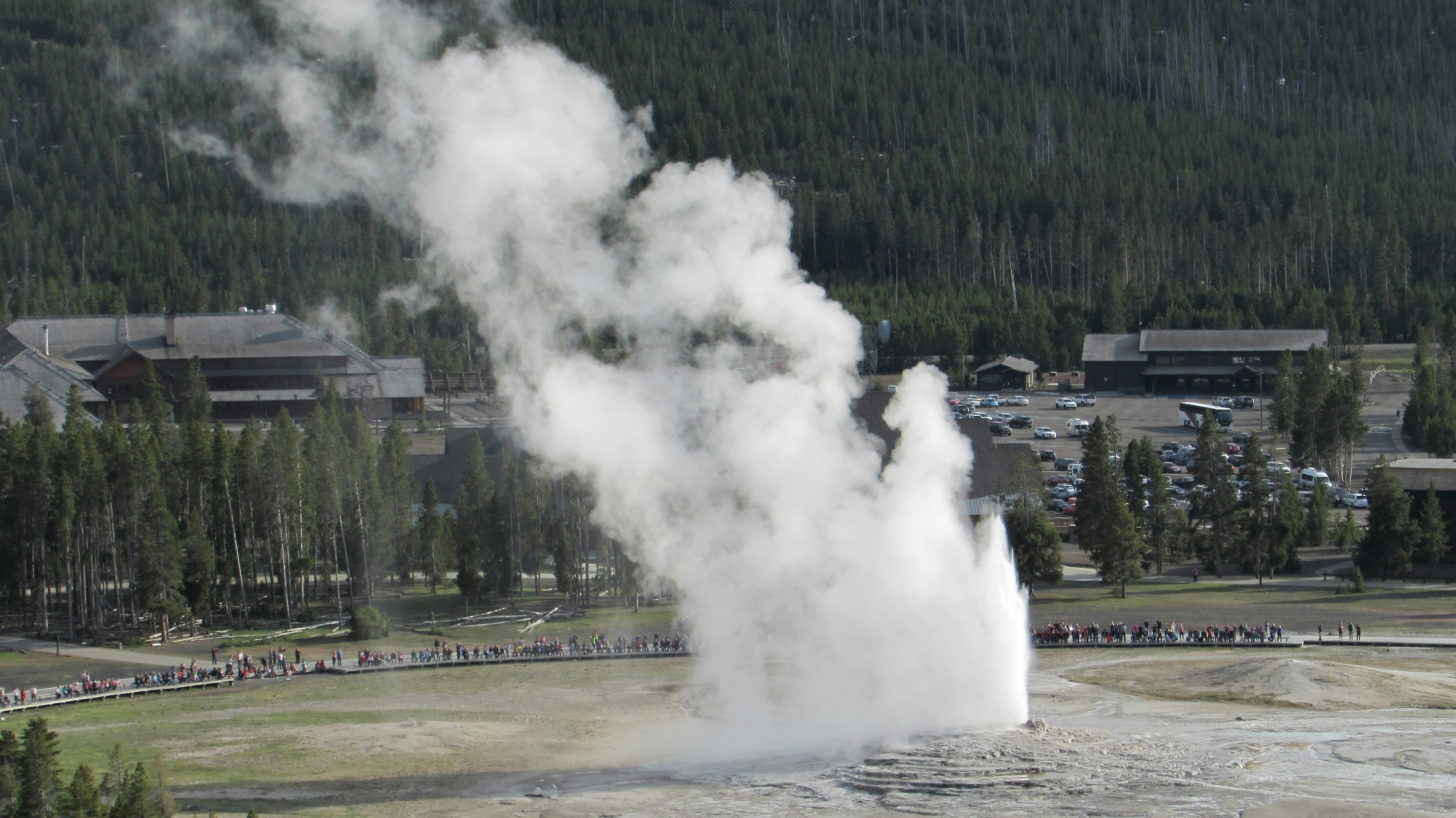 Geyser eruption sends a towering plume into the sky as crowds line a wooden boardwalk, with park buildings and pine trees in the background.