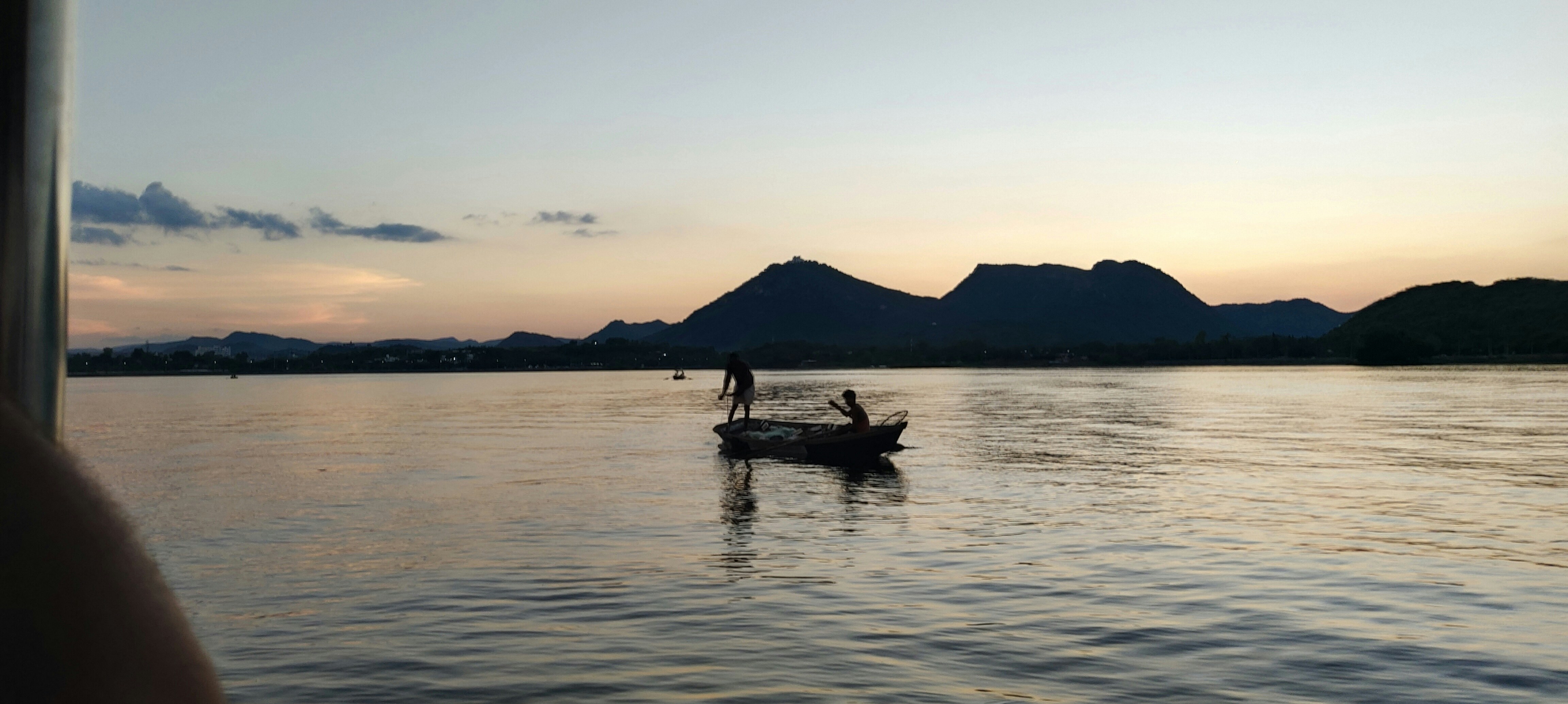 Two-person boat glides across calm water at sunset, silhouetted against distant mountains.