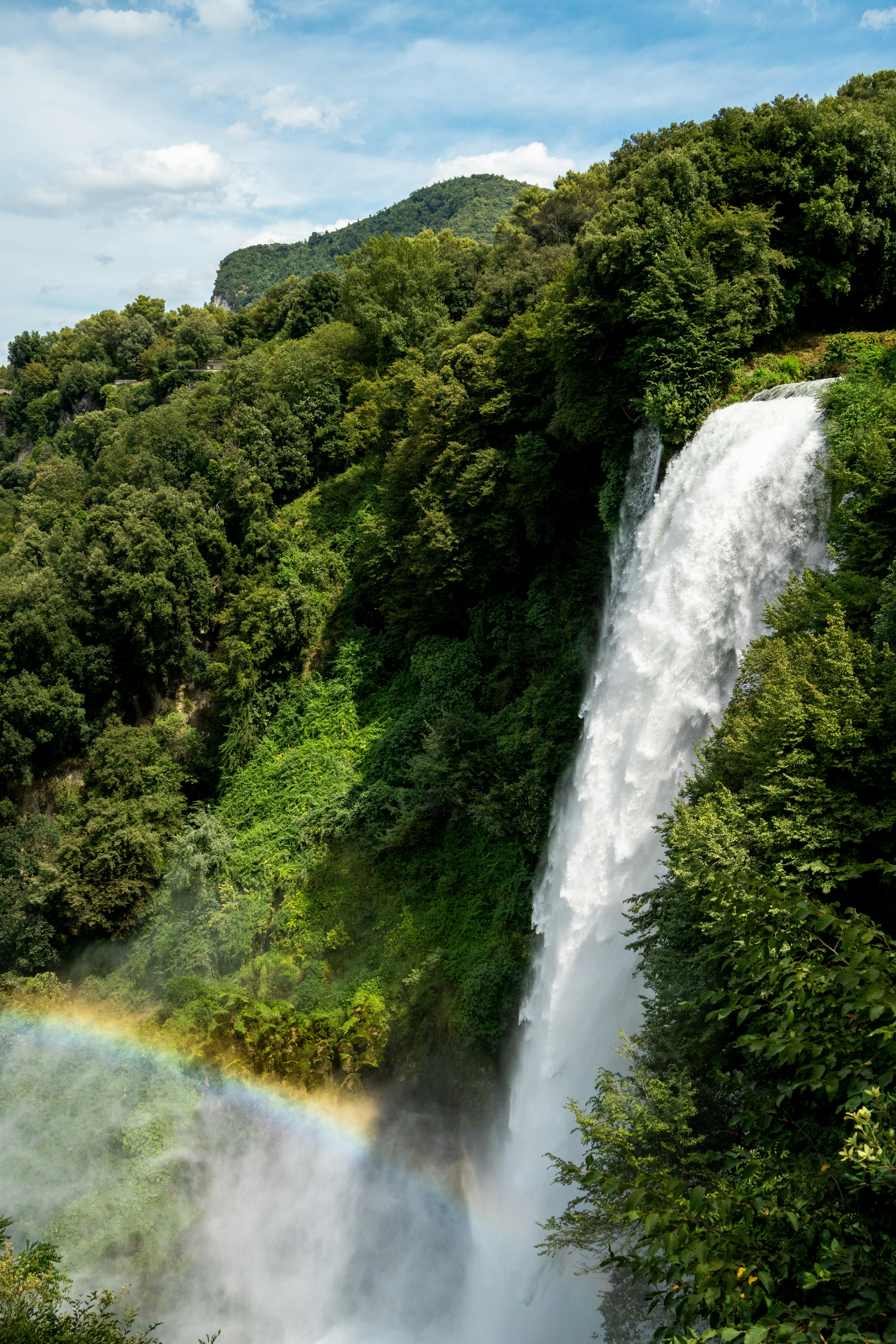 A majestic waterfall cascades down a lush green hillside, creating a vibrant rainbow at its base under a clear blue sky.