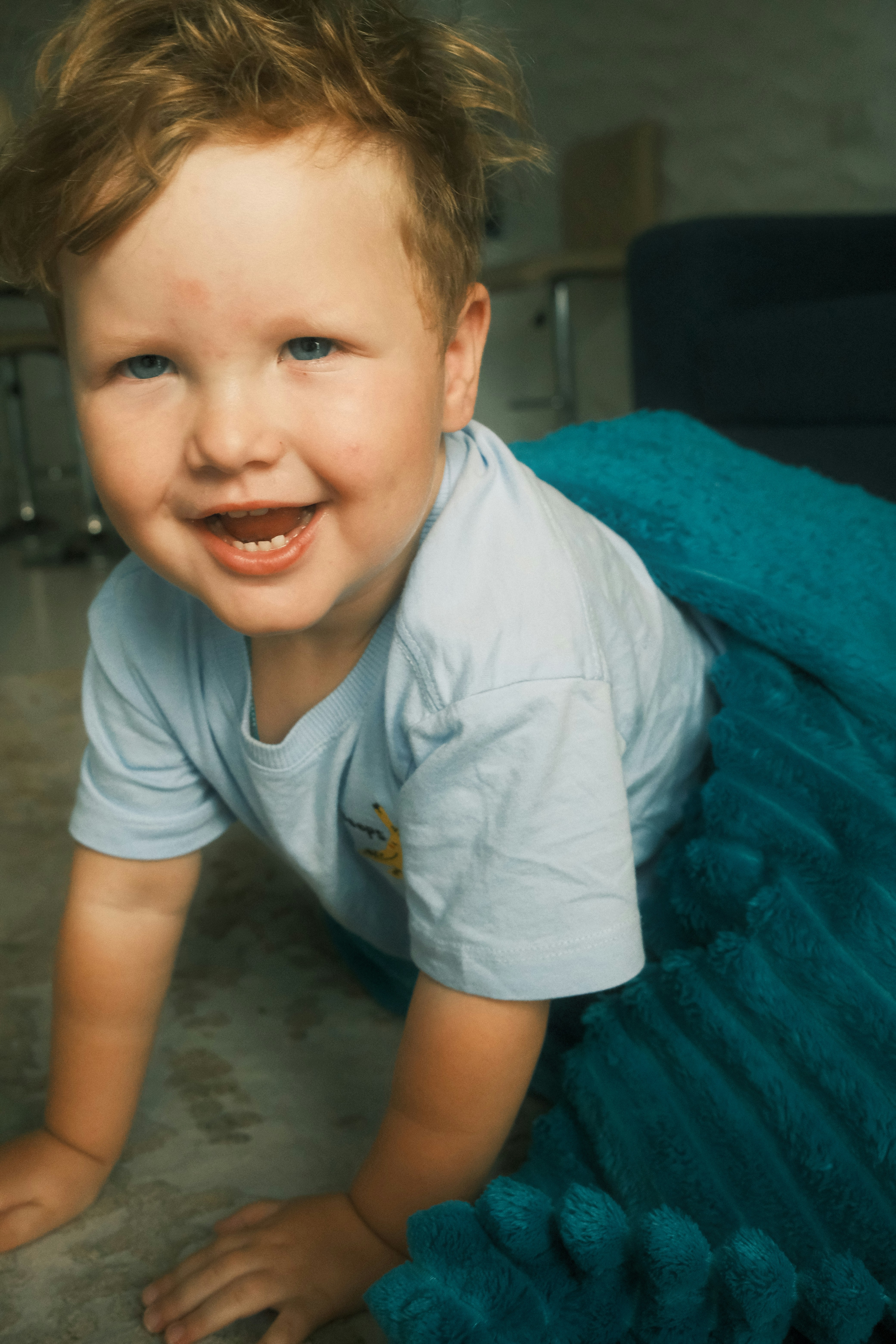 Close-up photograph of a smiling toddler crawling on a tiled floor, with a teal blanket behind.