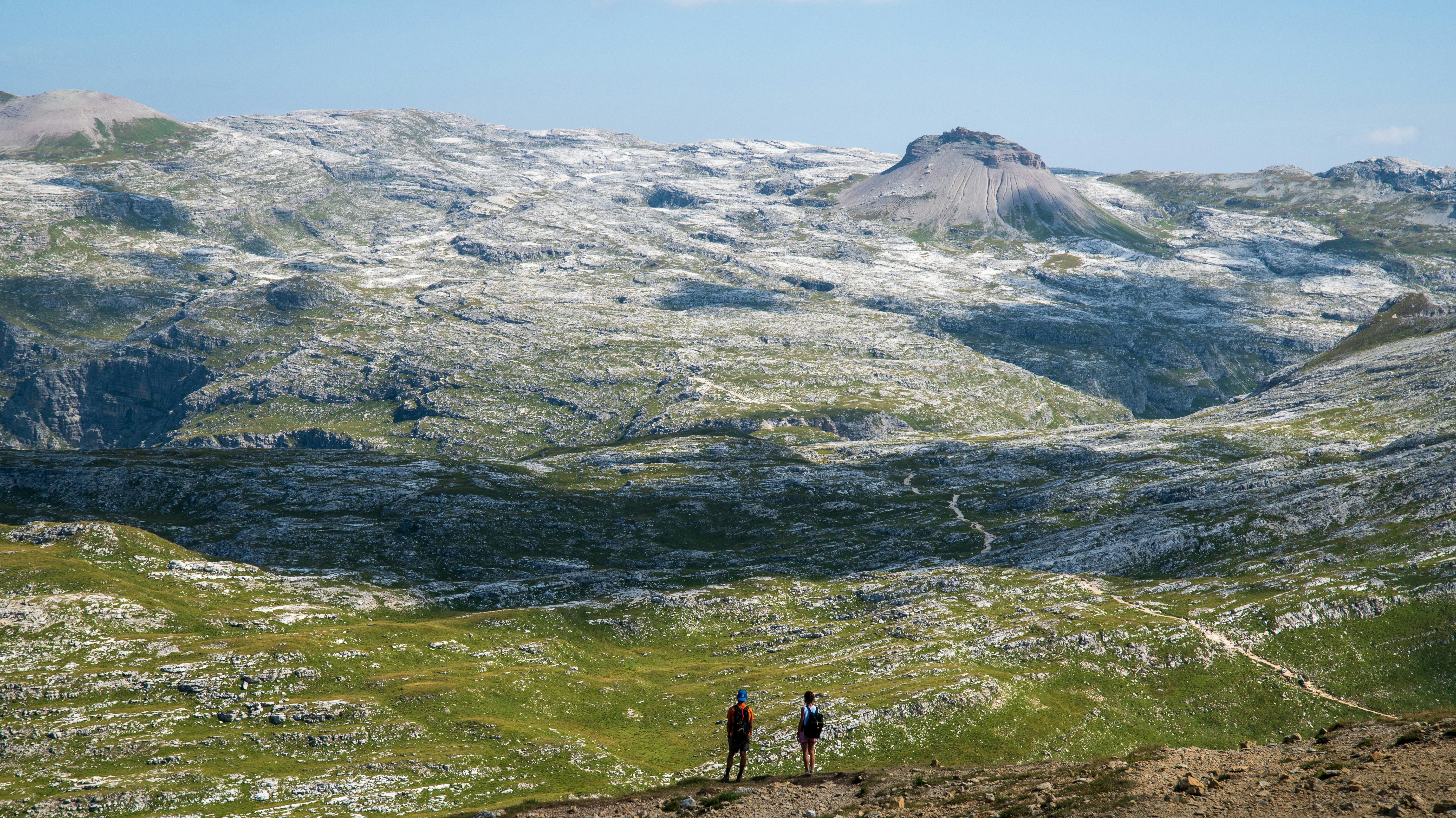 Two hikers traverse a sprawling, rocky landscape with snow-dusted peaks under a clear blue sky.