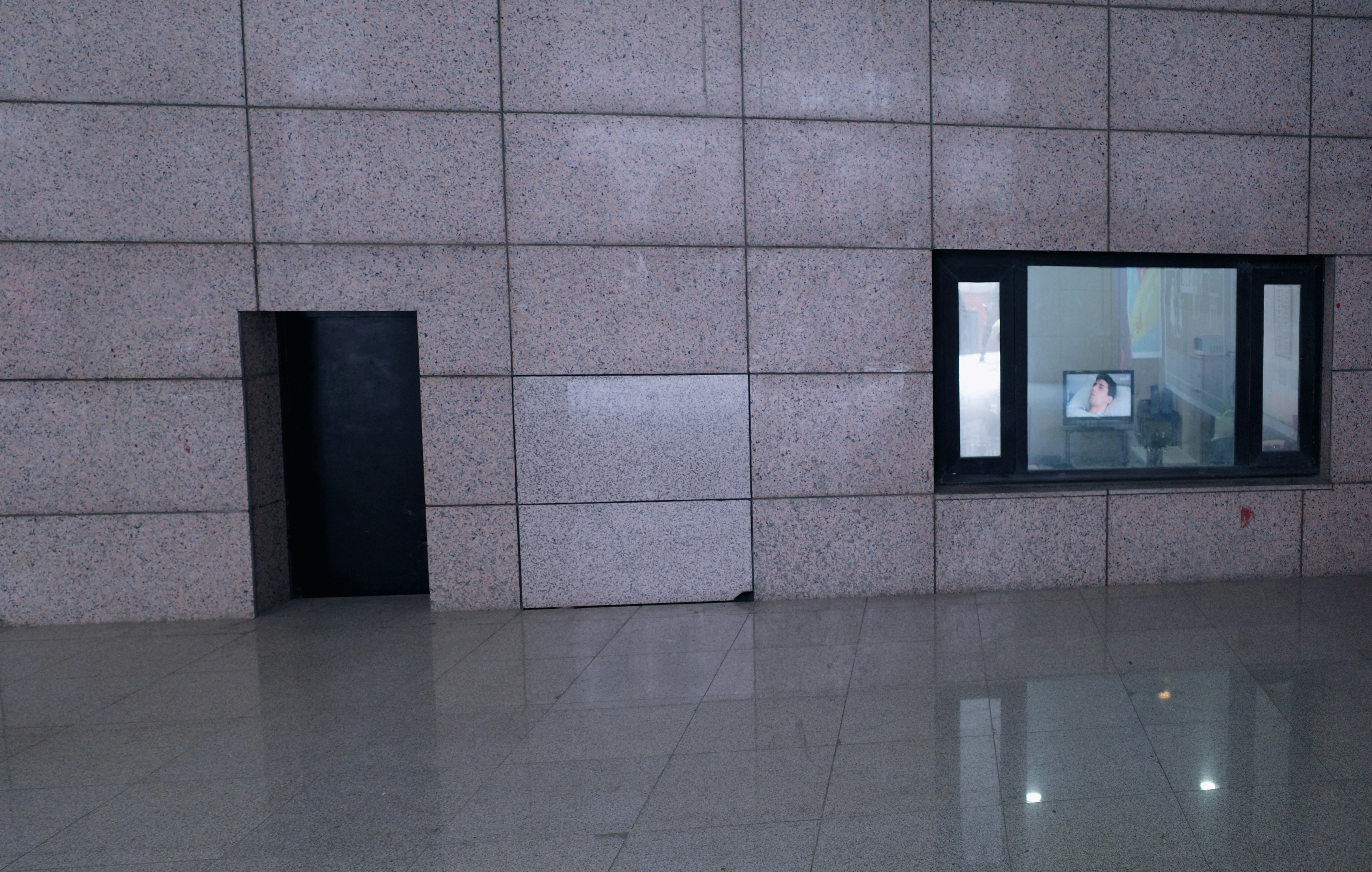 Minimalist interior scene featuring a textured tiled wall, a dark doorway on the left, and a window on the right displaying a monitor with a person.
