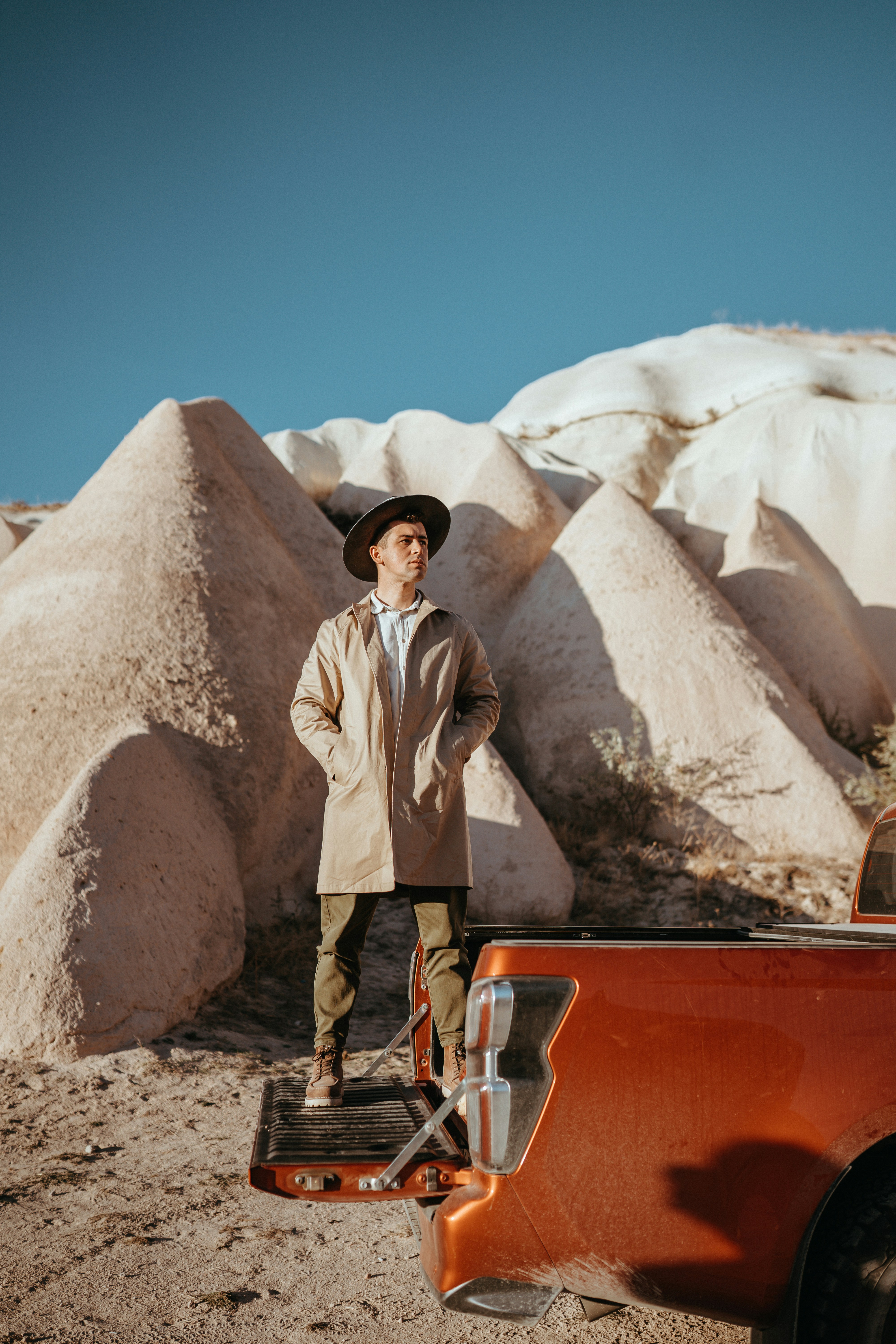 A man in a beige coat and hat stands confidently beside a vintage orange pickup truck, with unique rock formations in the background. The scene captures a sense of adventure and exploration.