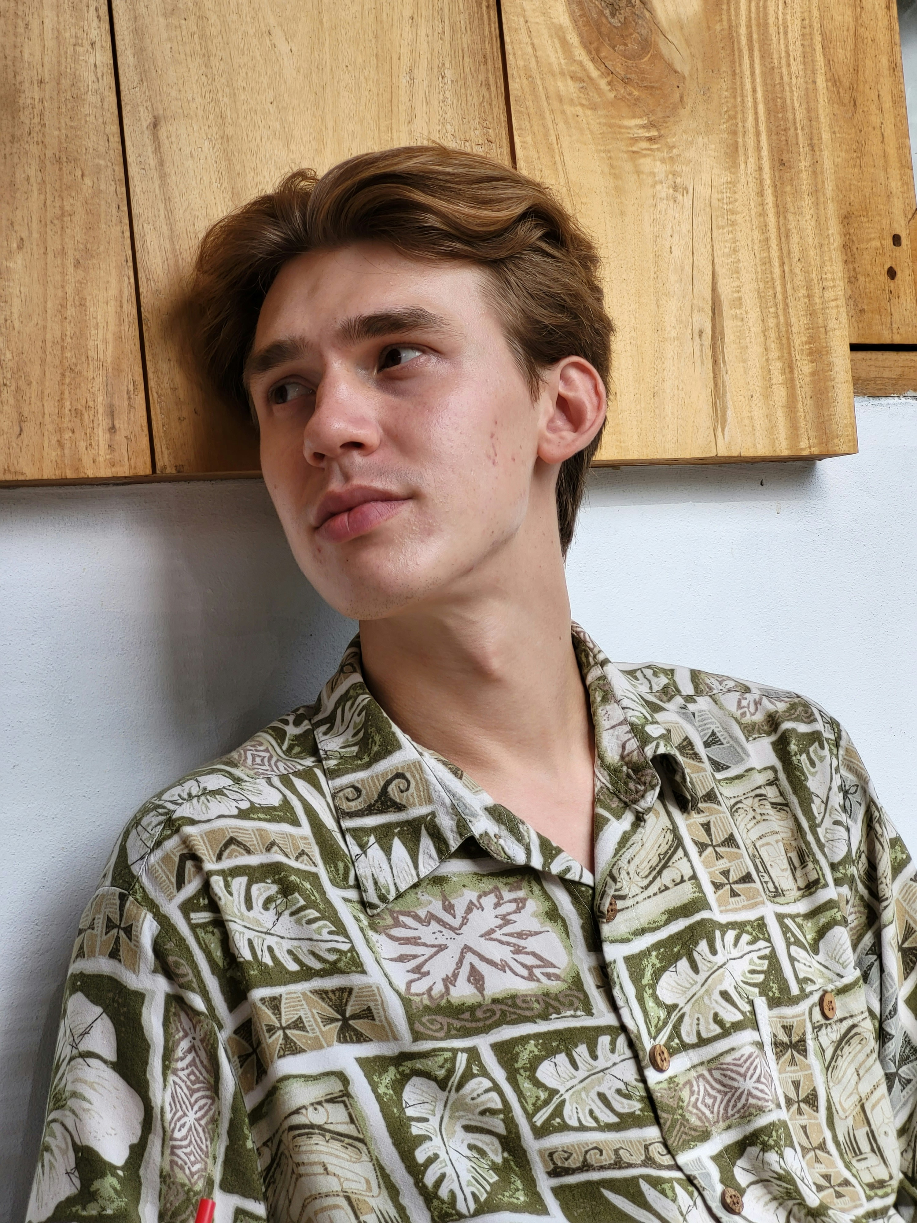 Portrait photo of a young man leaning against wooden panels, wearing a tropical-pattern shirt.