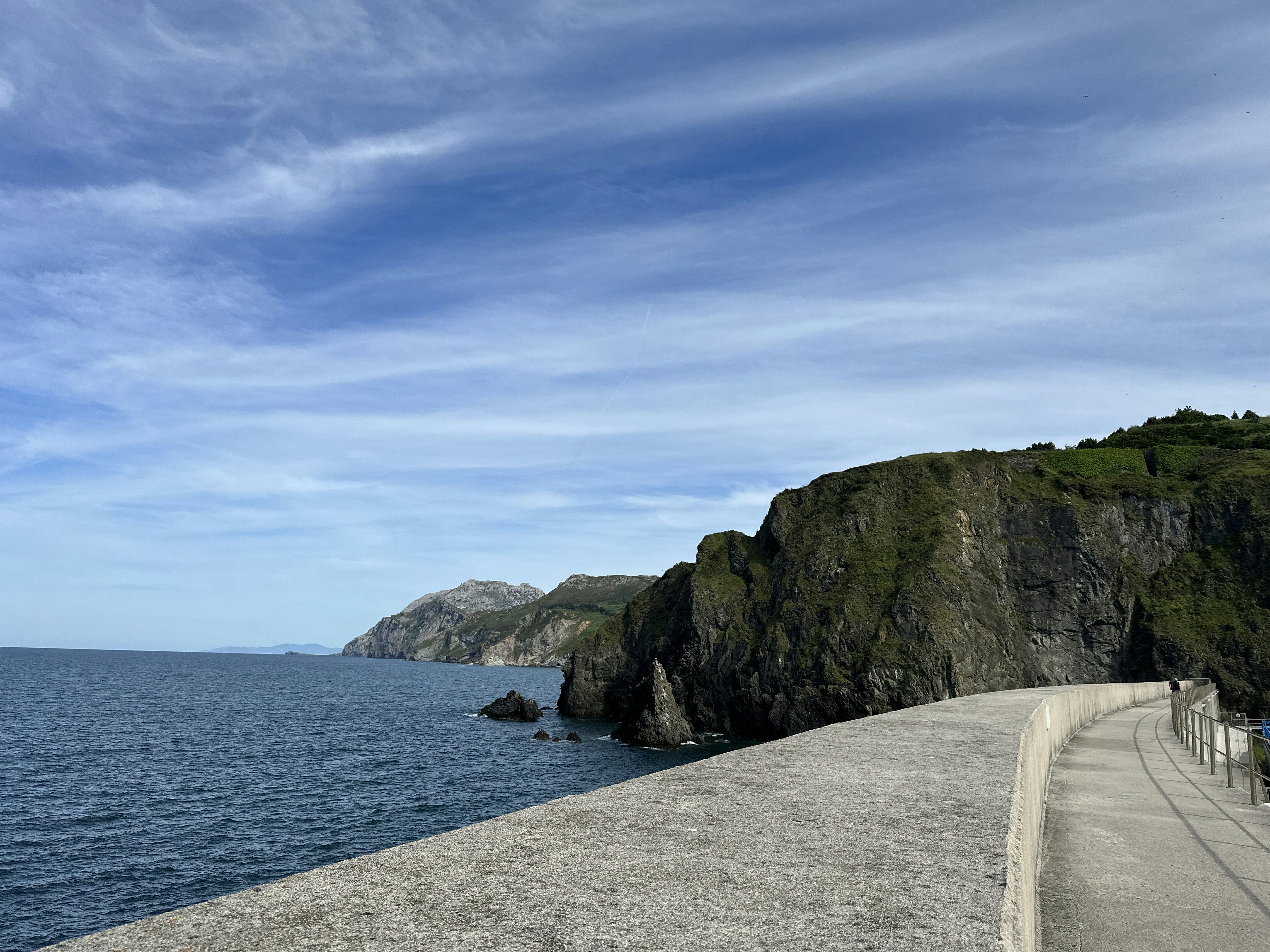 Curving seawall path leading to rugged cliffs under a vibrant blue sky.