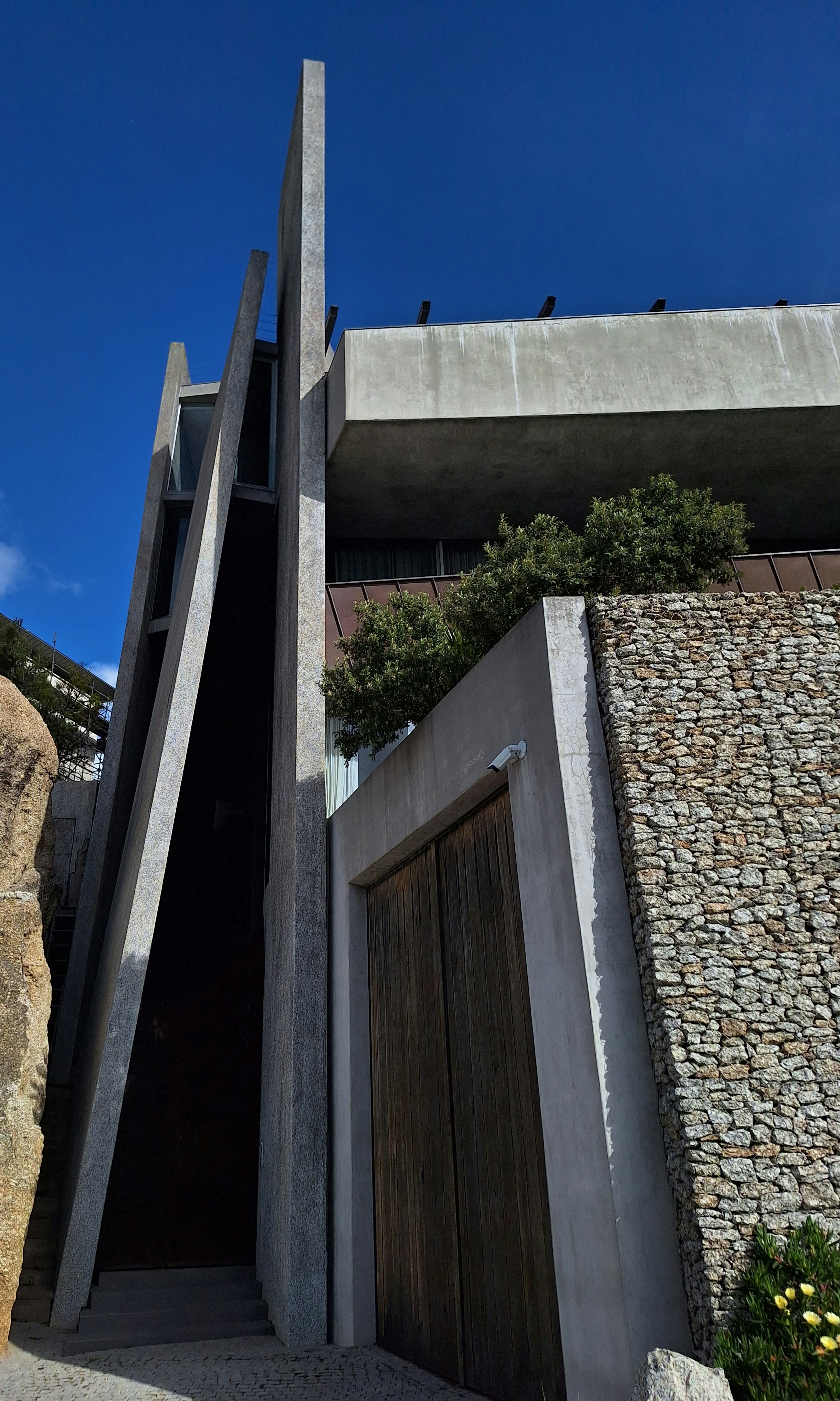 Modern concrete fins rise beside a dark doorway, framed by a stone wall and greenery, under a clear blue sky. The composition emphasizes geometric forms and material textures of contemporary architecture.
