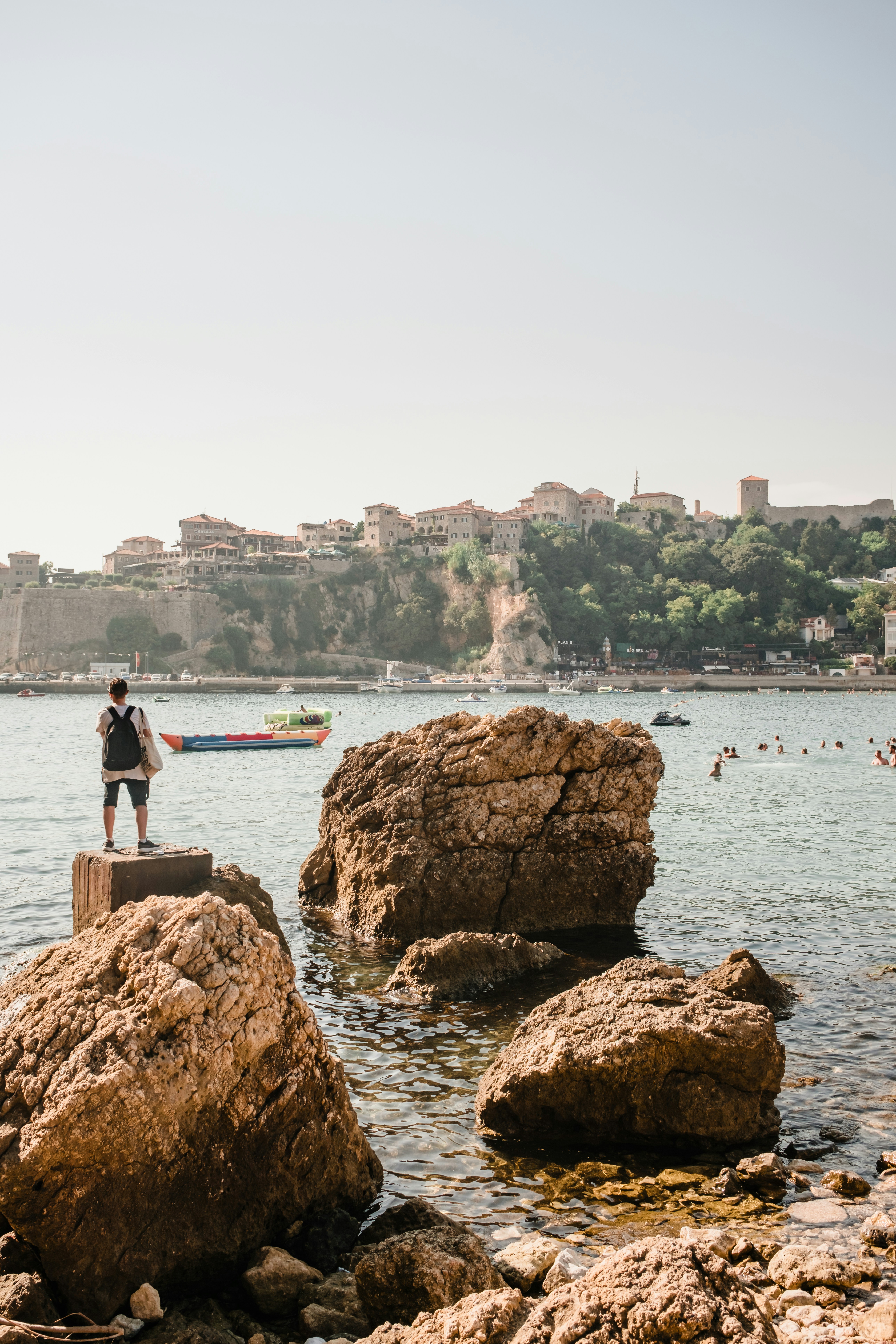 A solitary figure stands on a rocky outcrop, gazing across a tranquil sea towards a picturesque coastal town. The scene captures the essence of summer leisure.