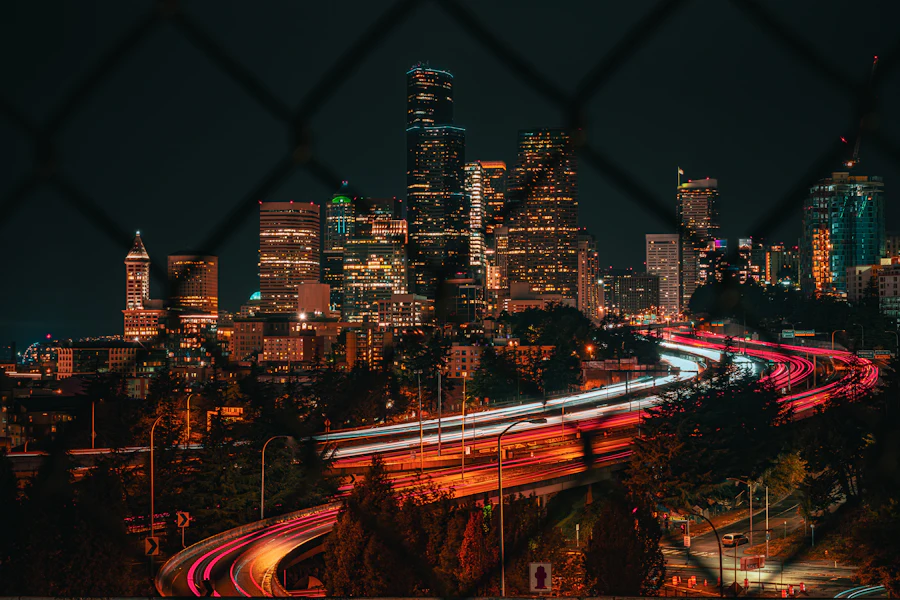 Illuminated downtown skyline at night with sweeping red and white highway light trails in the foreground. Dark sky and silhouetted trees frame the cityscape.