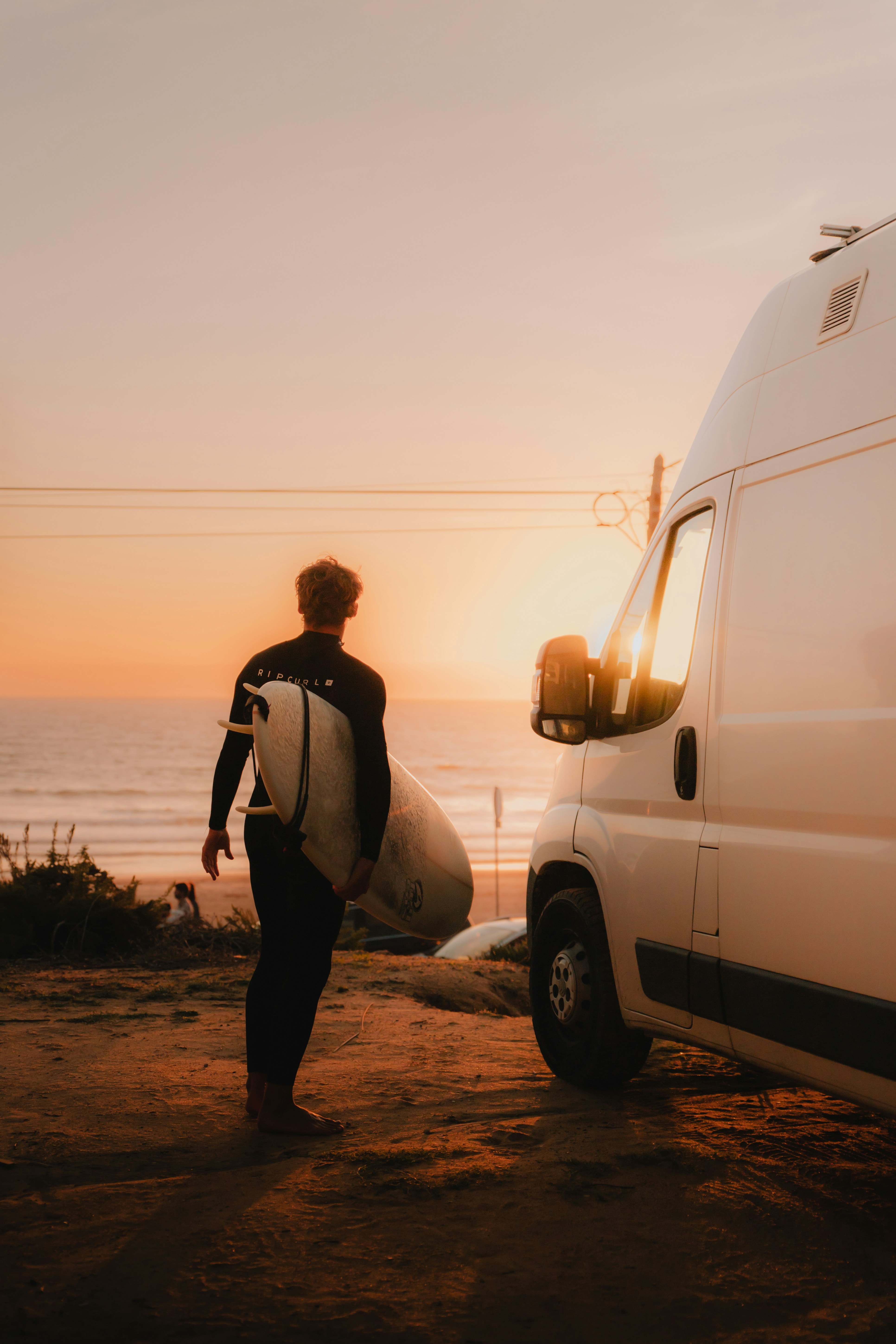 A surfer carrying a board walks toward a white van parked on the beach at sunset. The warm glow from the setting sun reflects off the water and the vehicle, highlighting the scene.