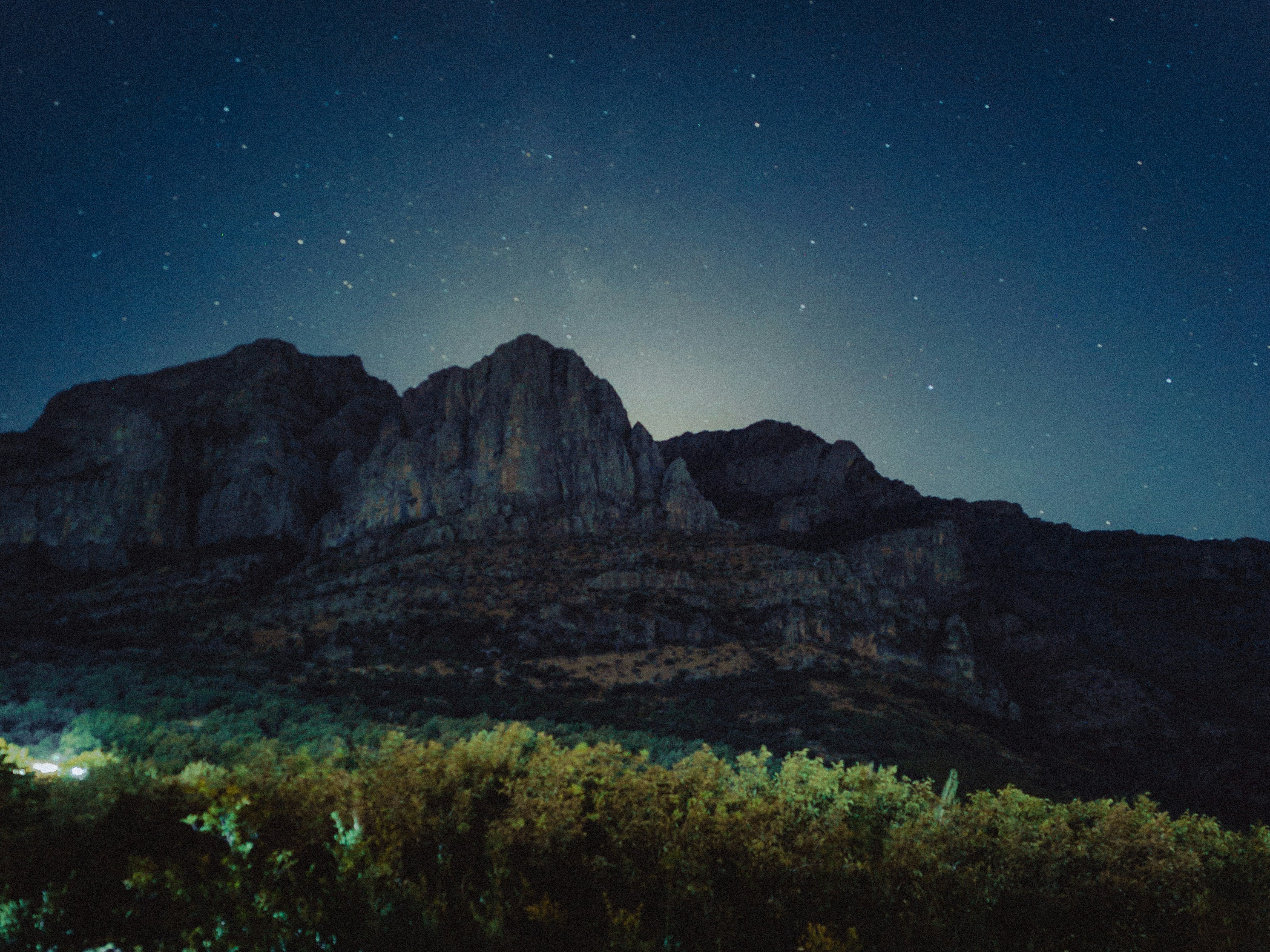 Star-filled night sky above a silhouetted mountain range with a soft glow on the horizon.