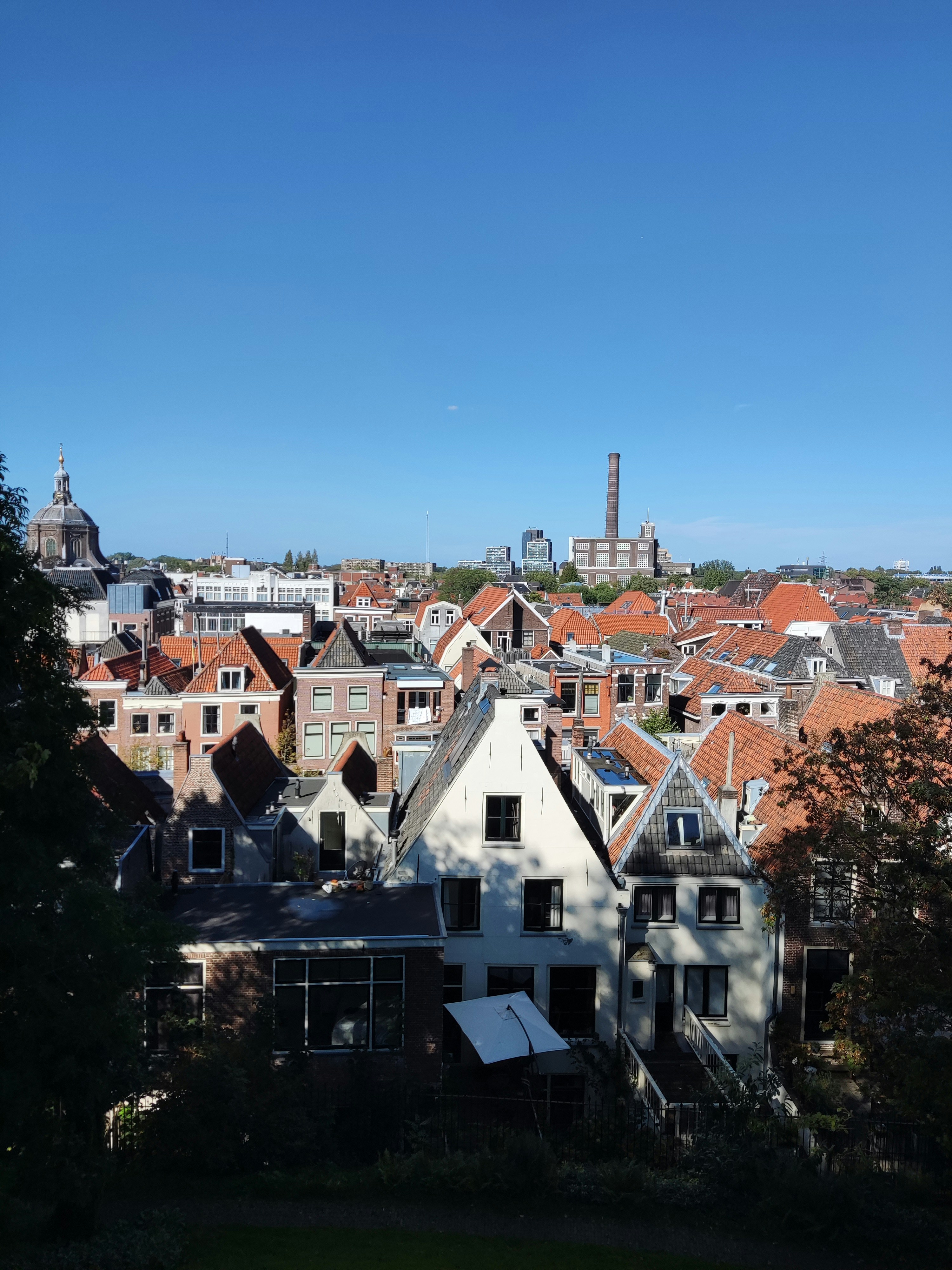 Cityscape of red-tiled rooftops and white gables fills the frame beneath a bright blue sky. A distant industrial stack rises on the horizon.