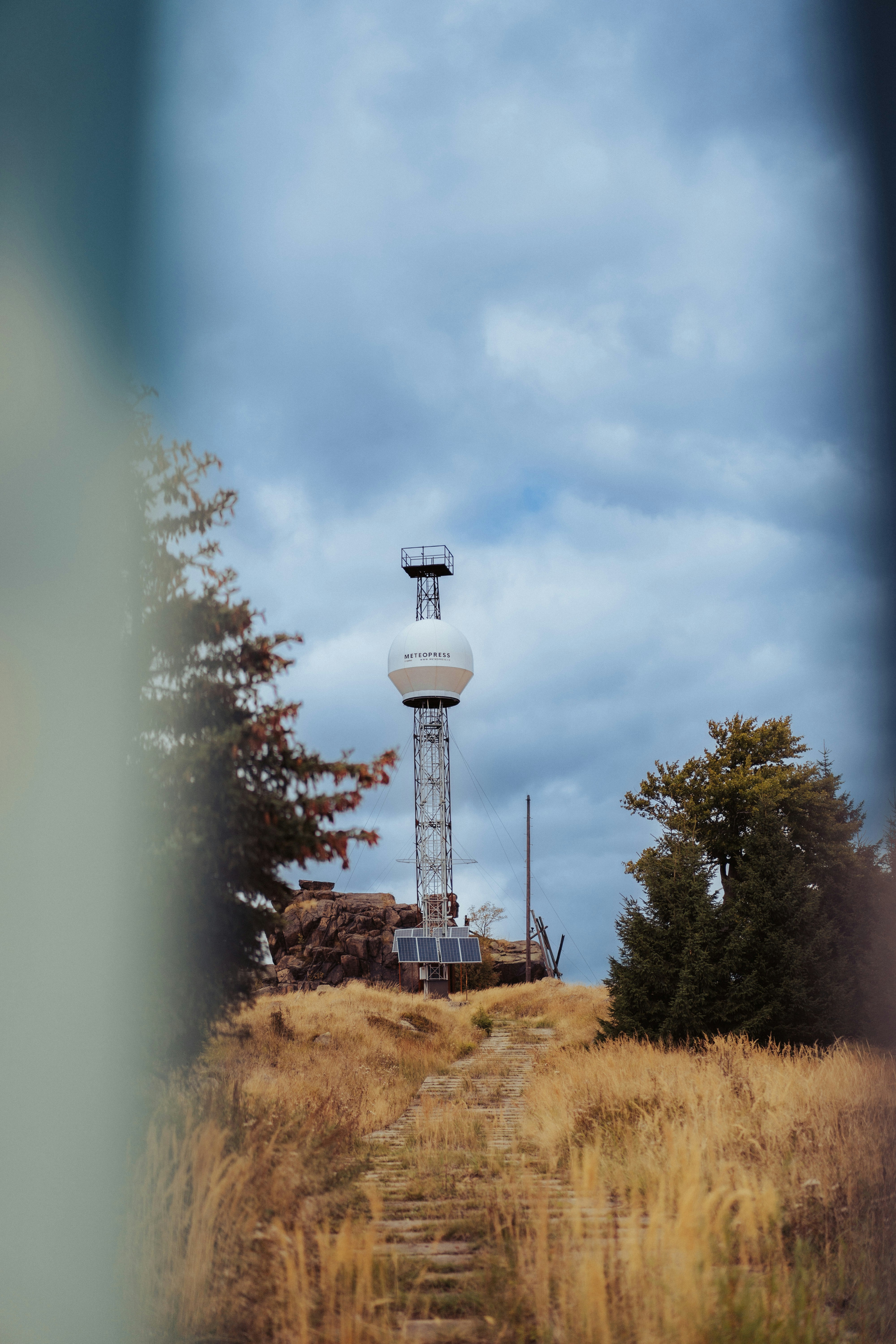 Communication tower standing tall amidst a grassy path and scattered trees under a cloudy sky.