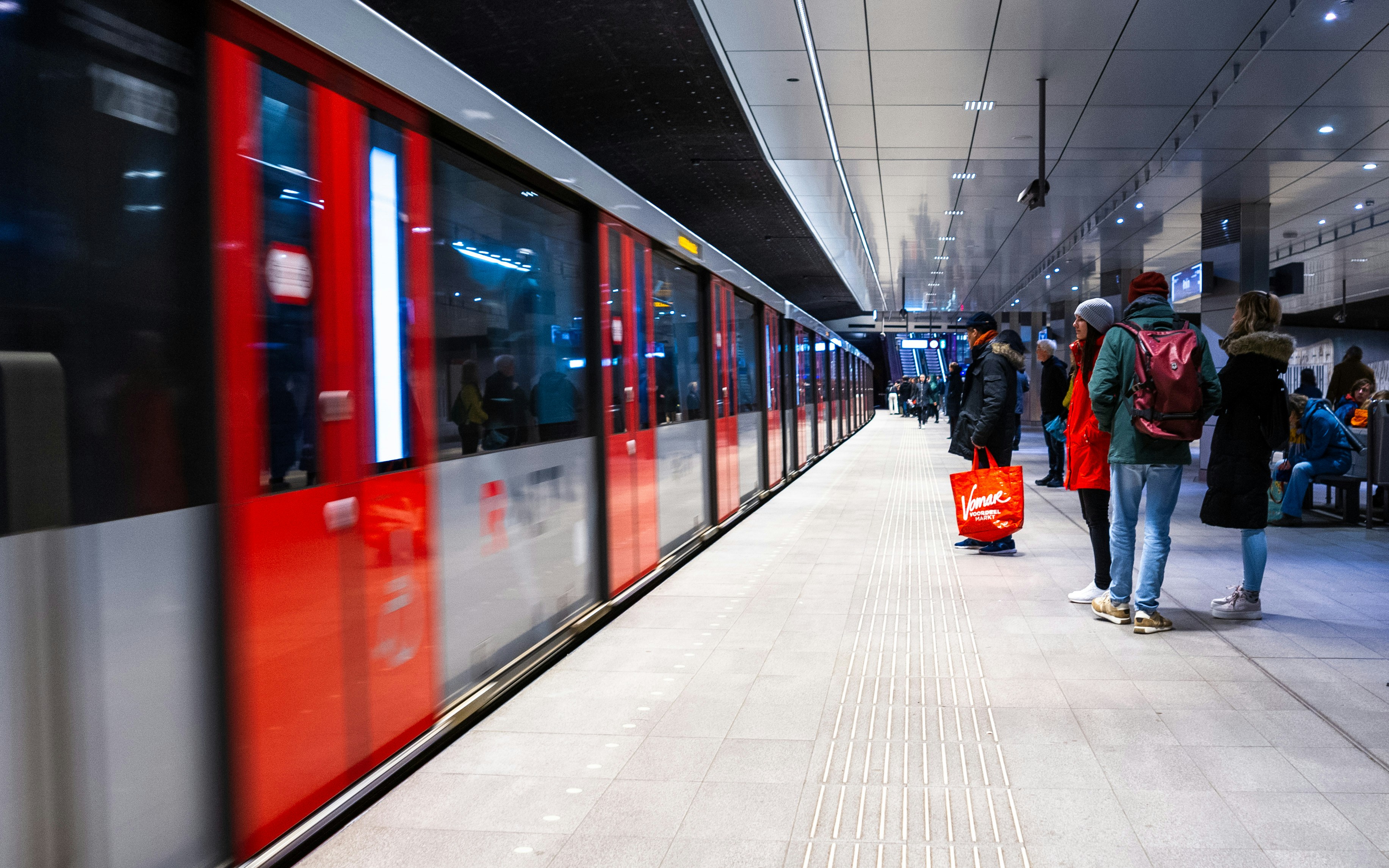 A bustling subway platform photograph showing a red train along the left and passengers waiting under bright overhead lighting.