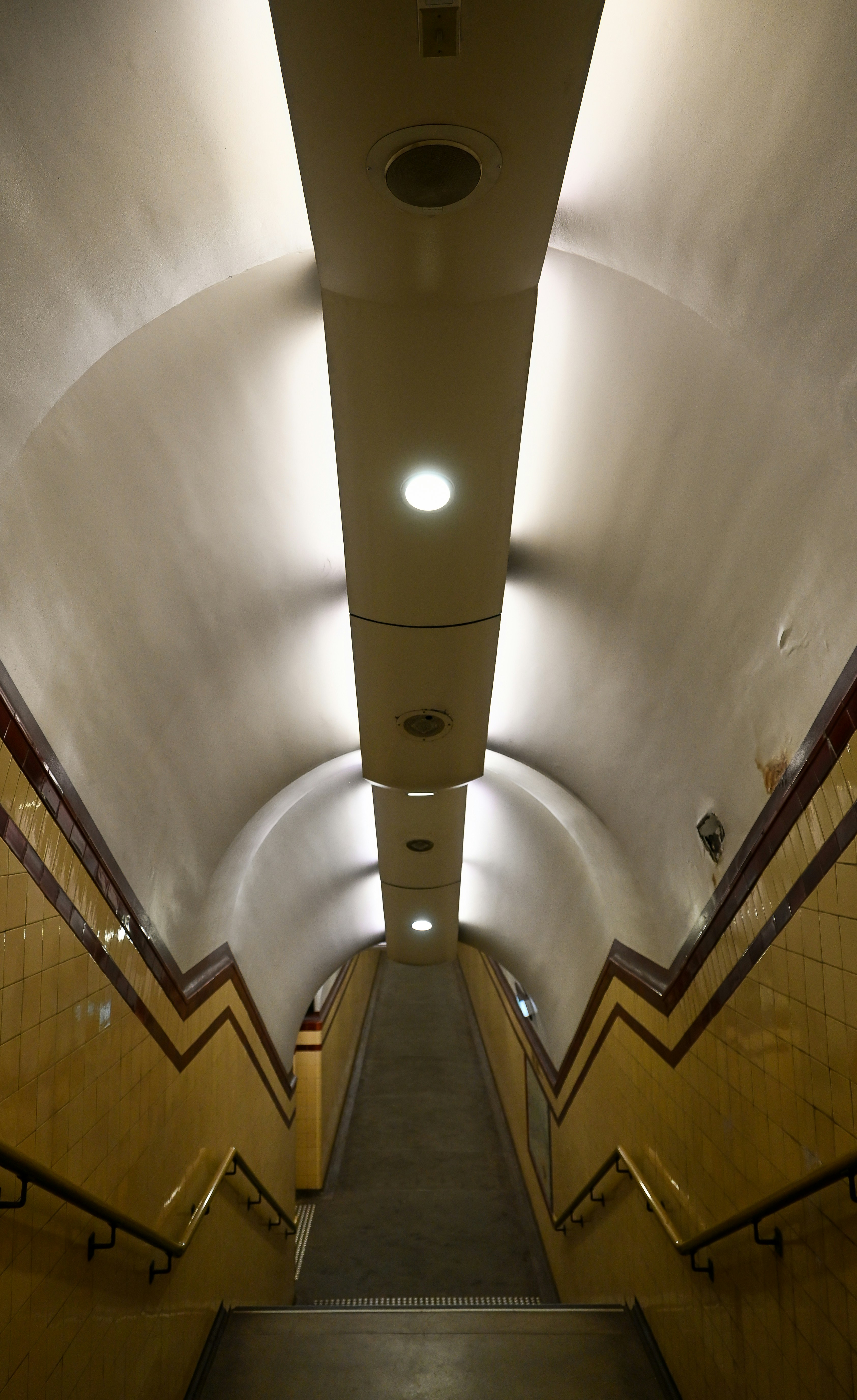 Curved tunnel with illuminated ceiling lights and yellow tiled walls leading downwards. The architectural design evokes a sense of history and depth.