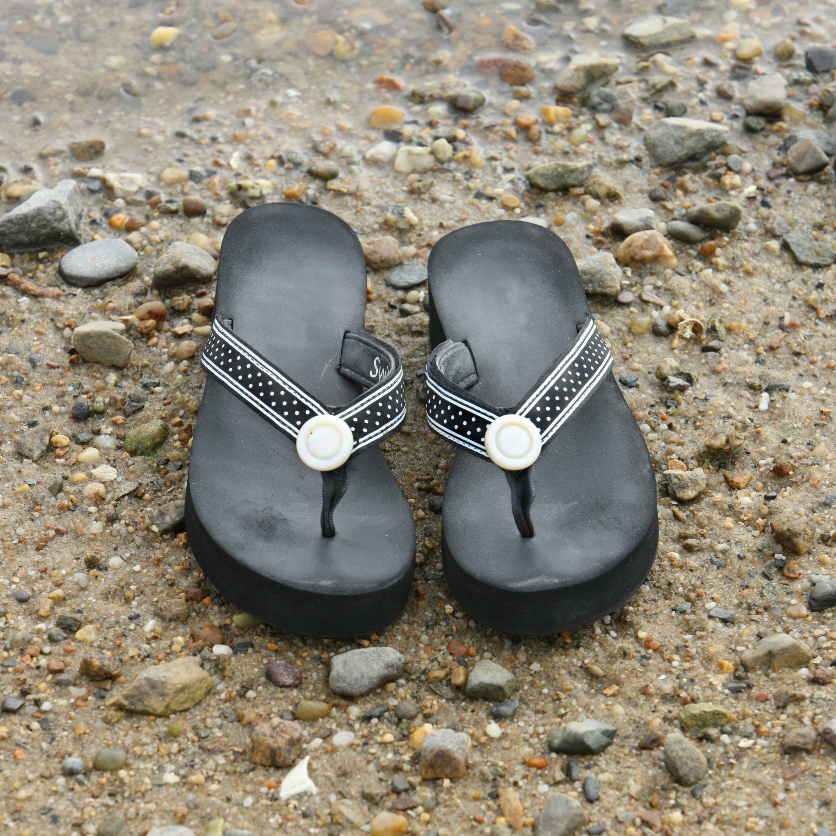 Black flip-flops resting on a sandy shore, surrounded by small pebbles and water. The scene evokes a sense of leisure and relaxation.