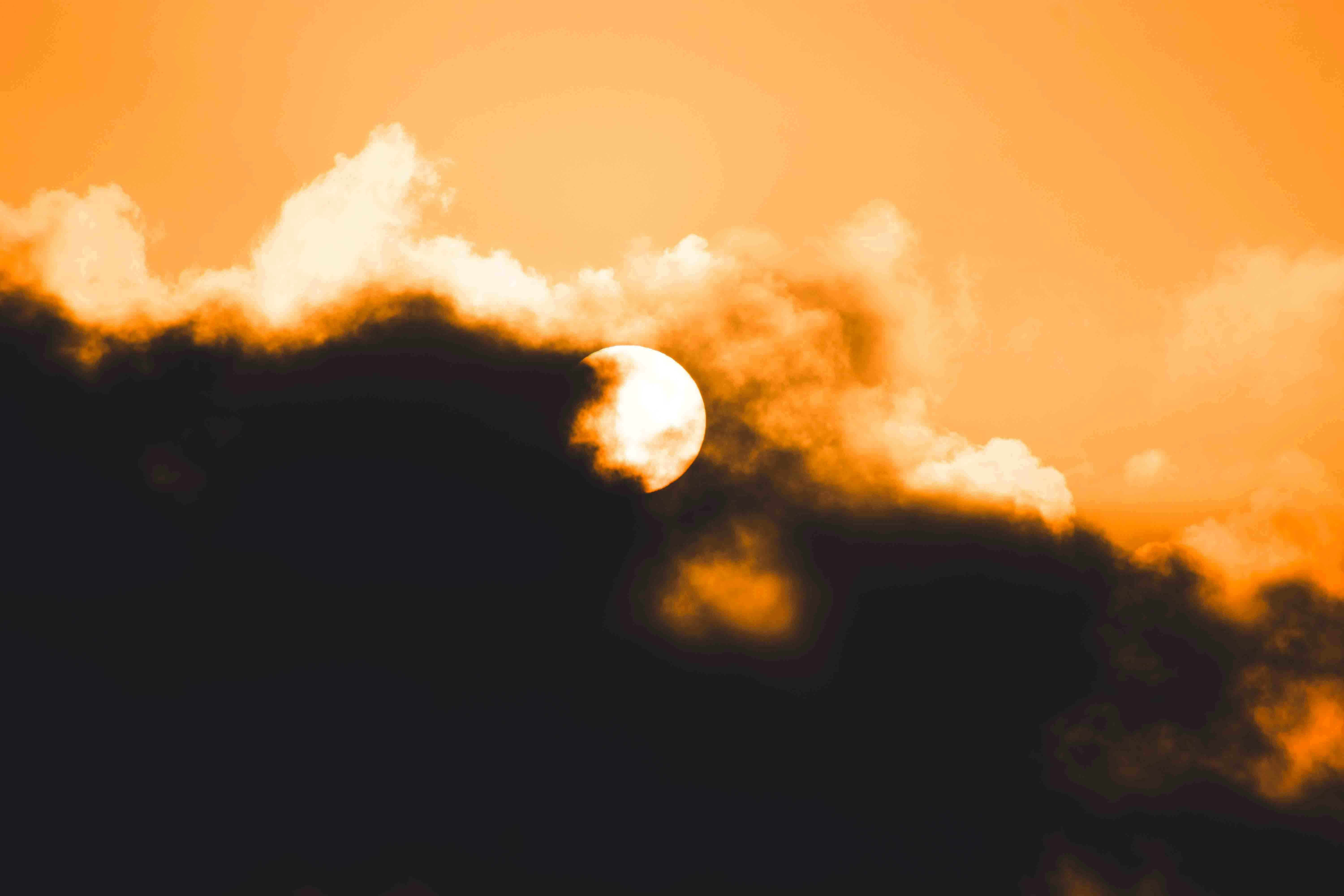Silhouetted plane traverses a vibrant orange sky with the moon partially obscured by clouds.