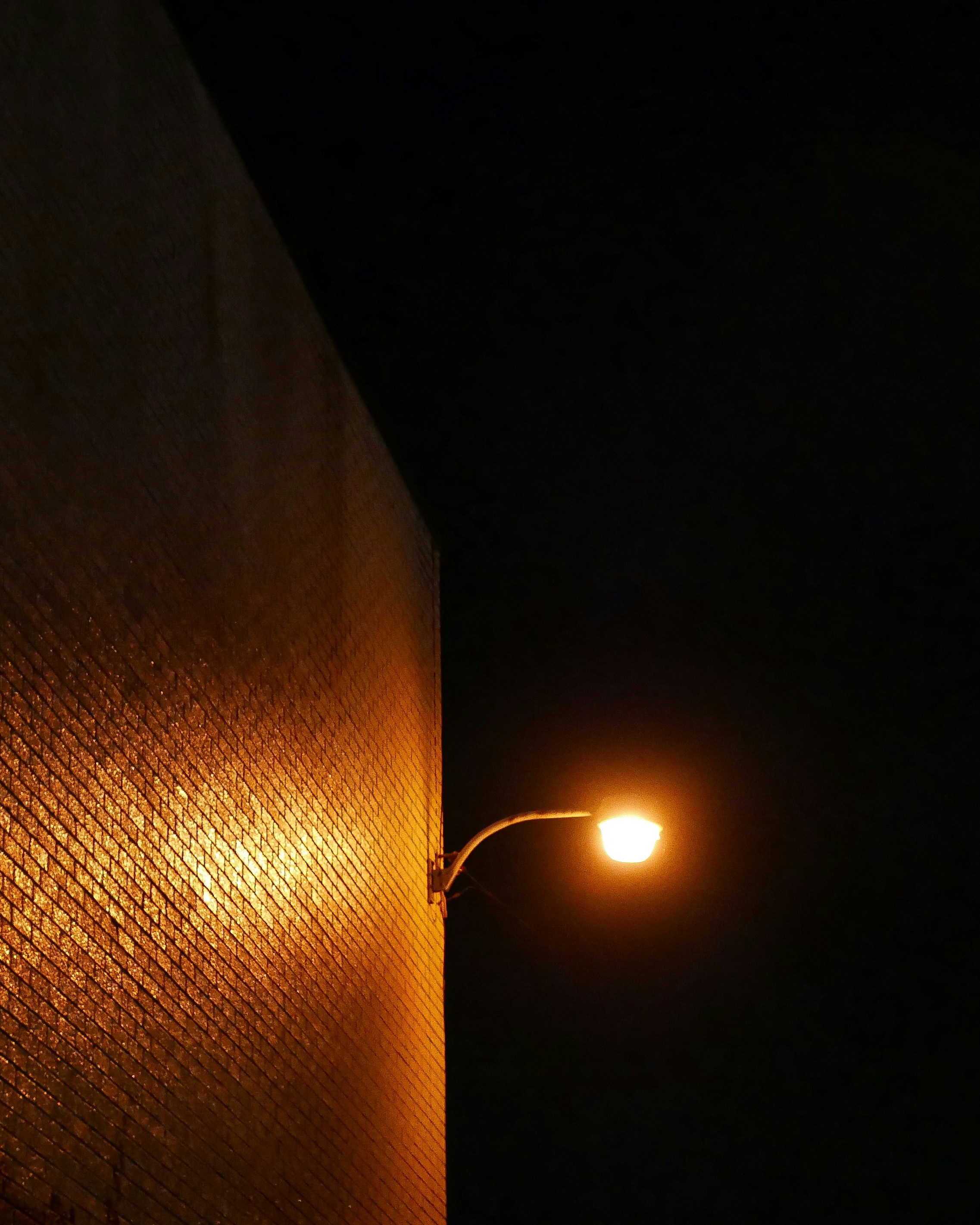 A solitary streetlight casts a warm glow on a textured brick wall at night, highlighting the interplay of light and shadow.