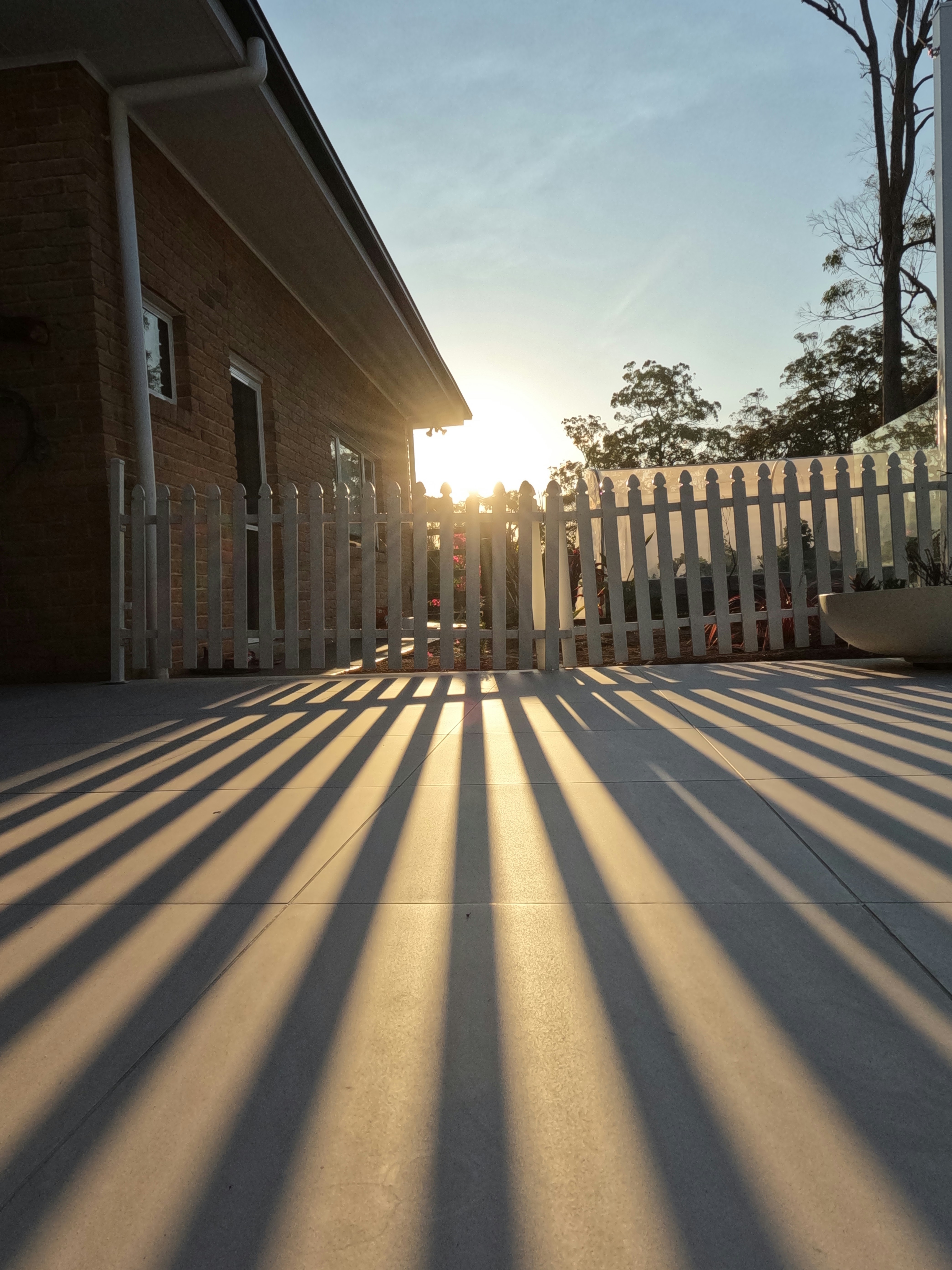 Sunlight floods a patio with long, dramatic shadows cast by a white picket fence, a brick house on the left, and potted plants on the right.