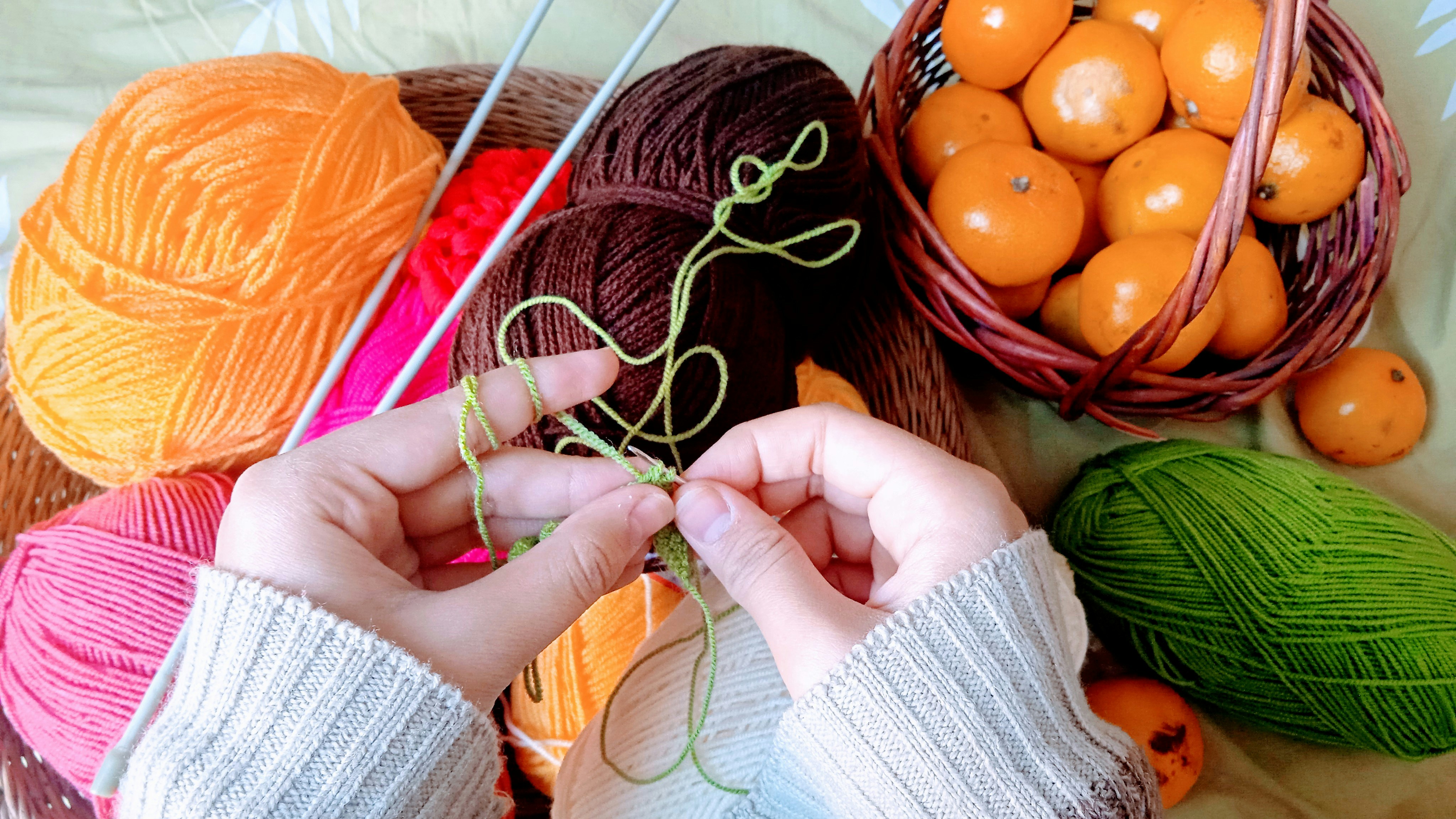 Close-up photograph of hands knitting with green yarn among colorful skeins, with a basket of oranges in the background.