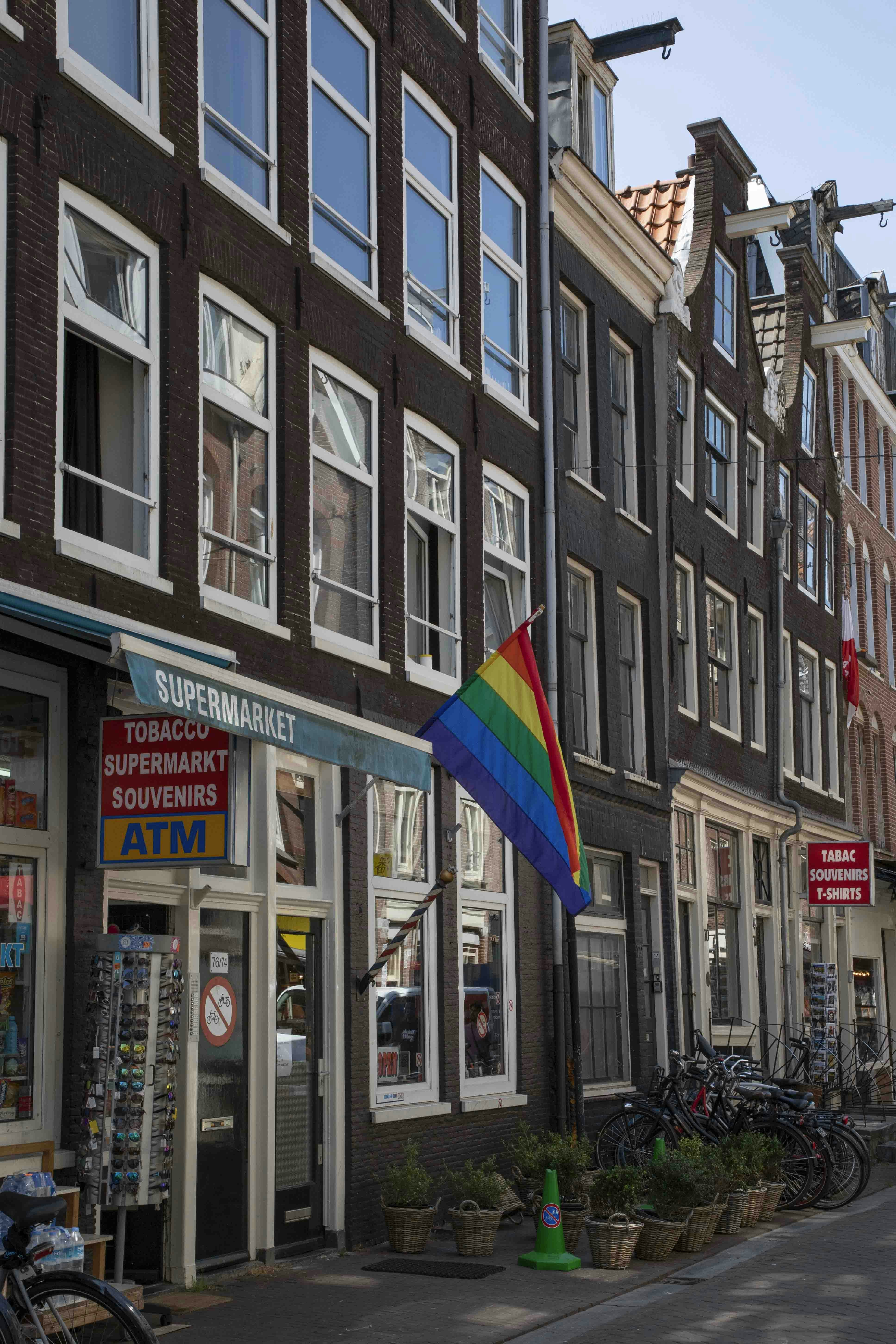 Colorful storefronts line a bustling street in Amsterdam, featuring a prominent rainbow flag and a variety of shops. The scene reflects local culture and community spirit.