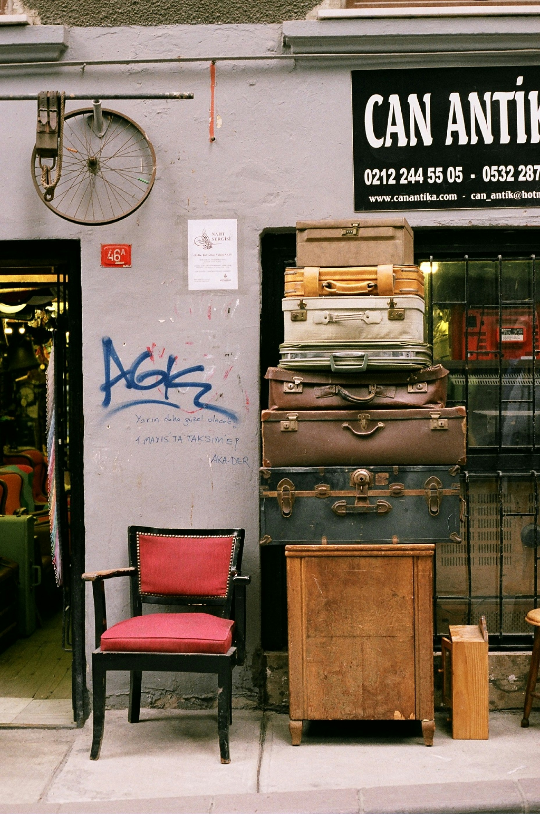 A street photograph of a stack of vintage suitcases beside a red chair in front of a graffiti-splashed shopfront.