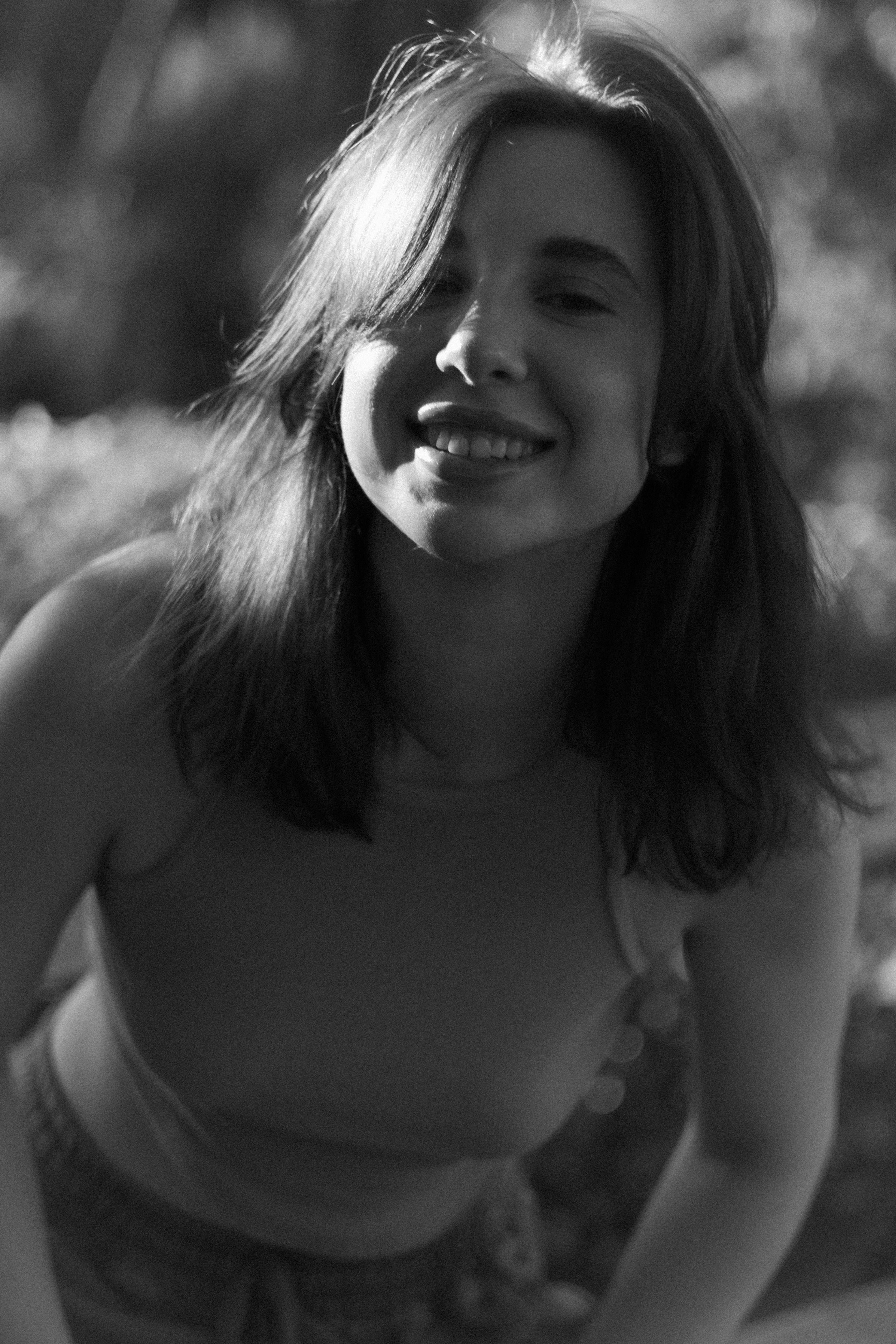 Monochrome portrait photograph of a smiling woman outdoors, with a shallow depth of field and soft bokeh that isolates the subject.