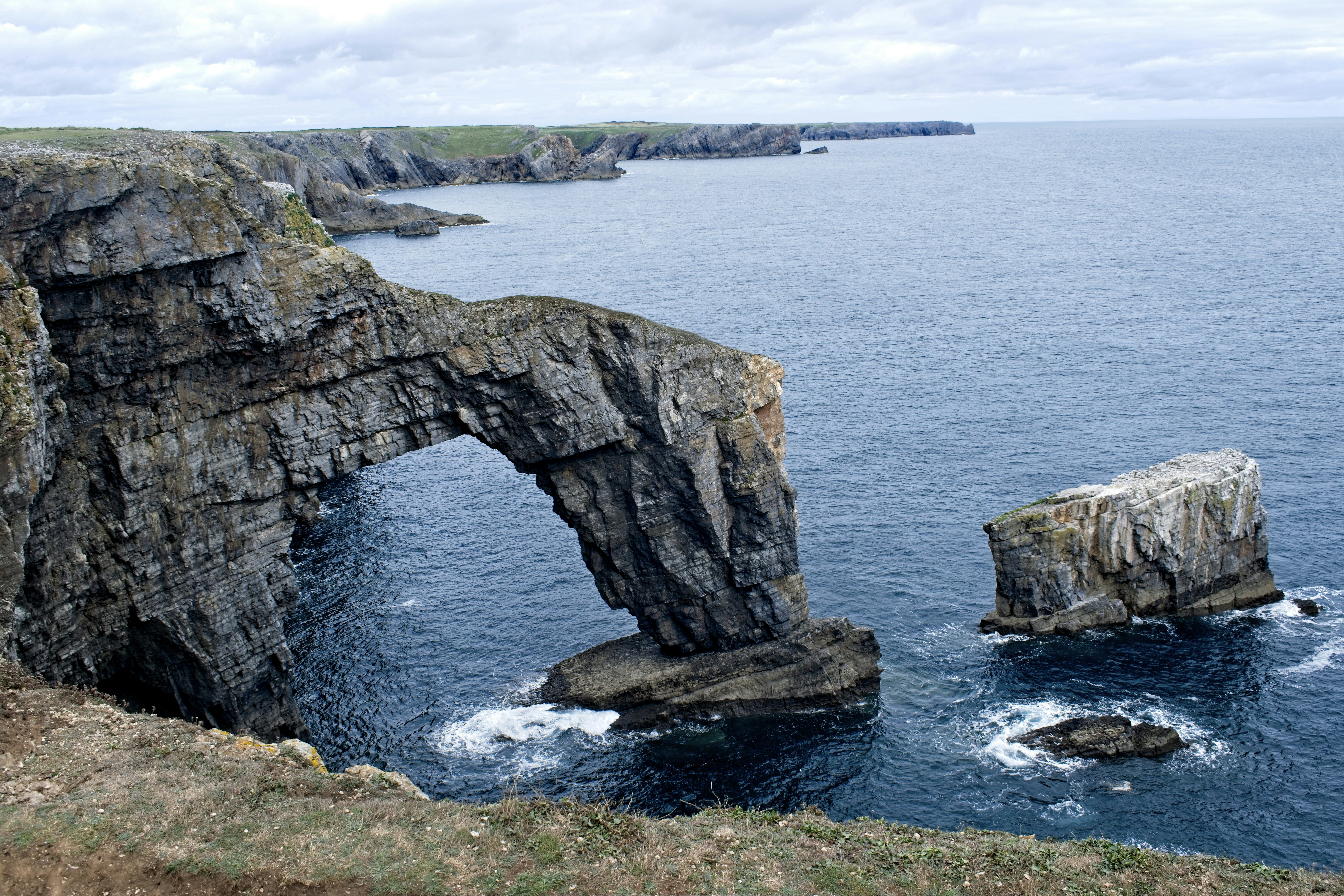 Natural rock arch over blue waves near a rugged coastline under a cloudy sky.