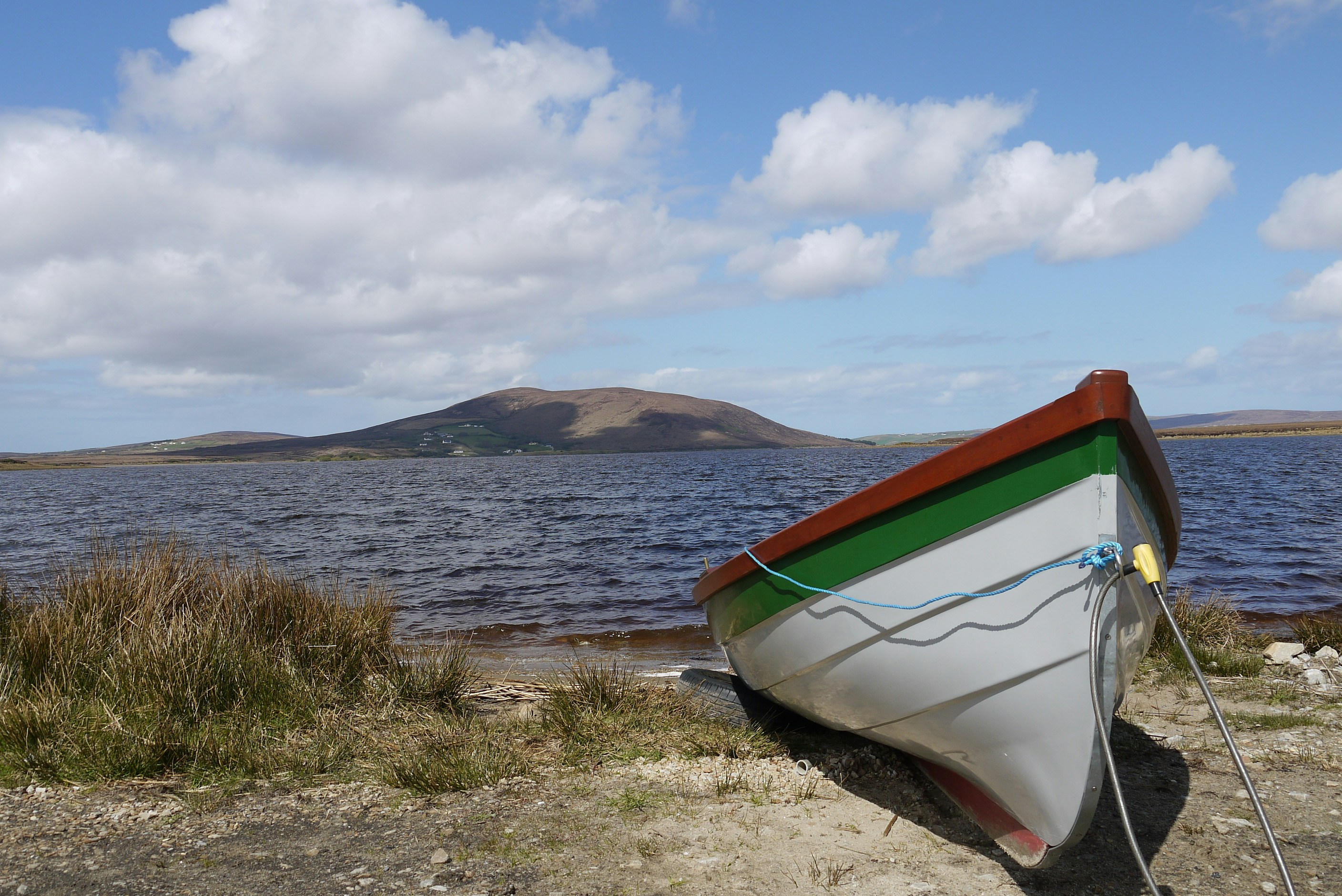 A small boat with a green interior rests on a pebbled shore; calm water stretches toward a distant island under a partly cloudy sky.