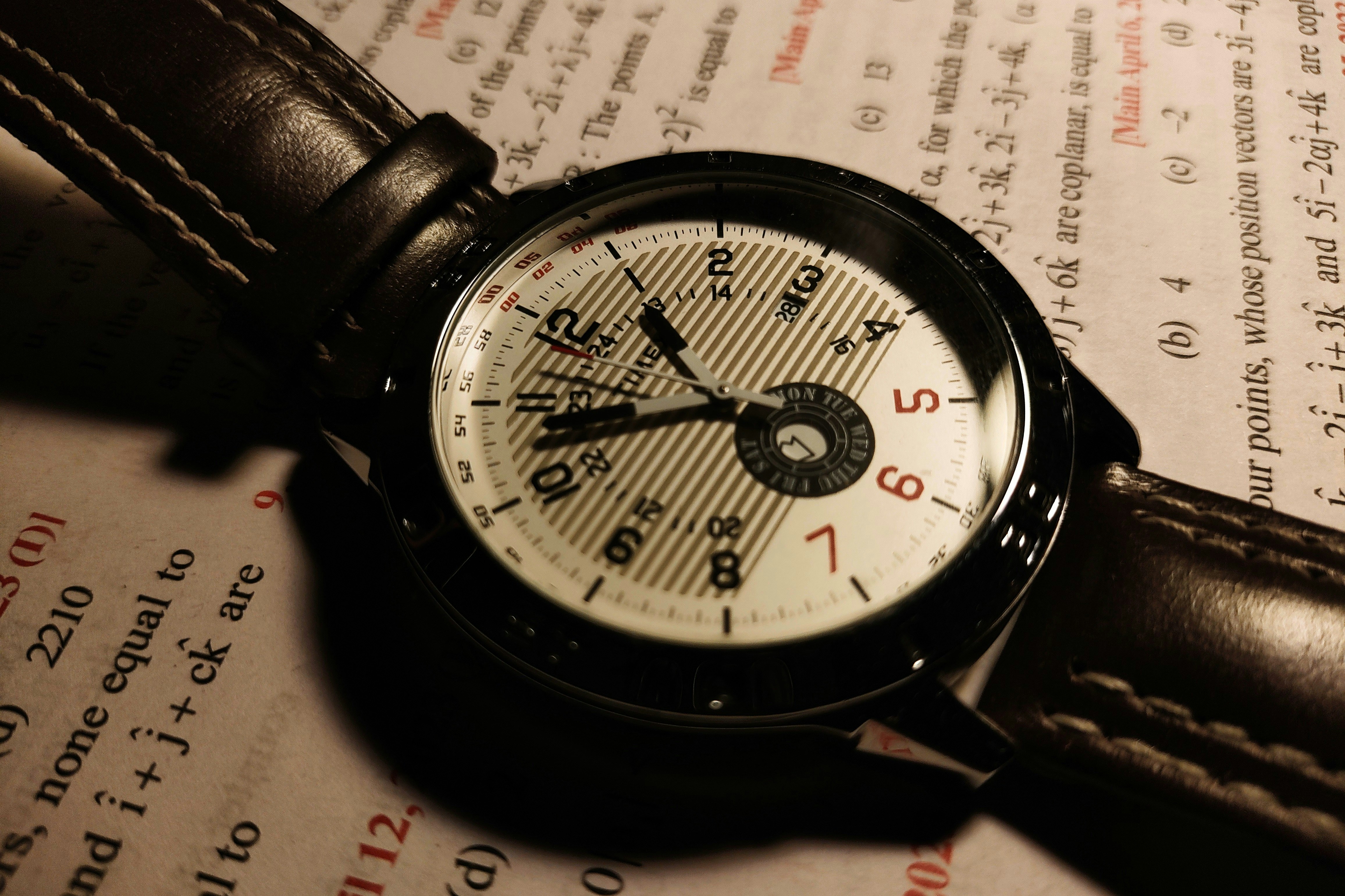 Analog wristwatch with a black case and brown leather strap rests on a page of mathematical text; the white dial features bold black numerals and subtle red accents.