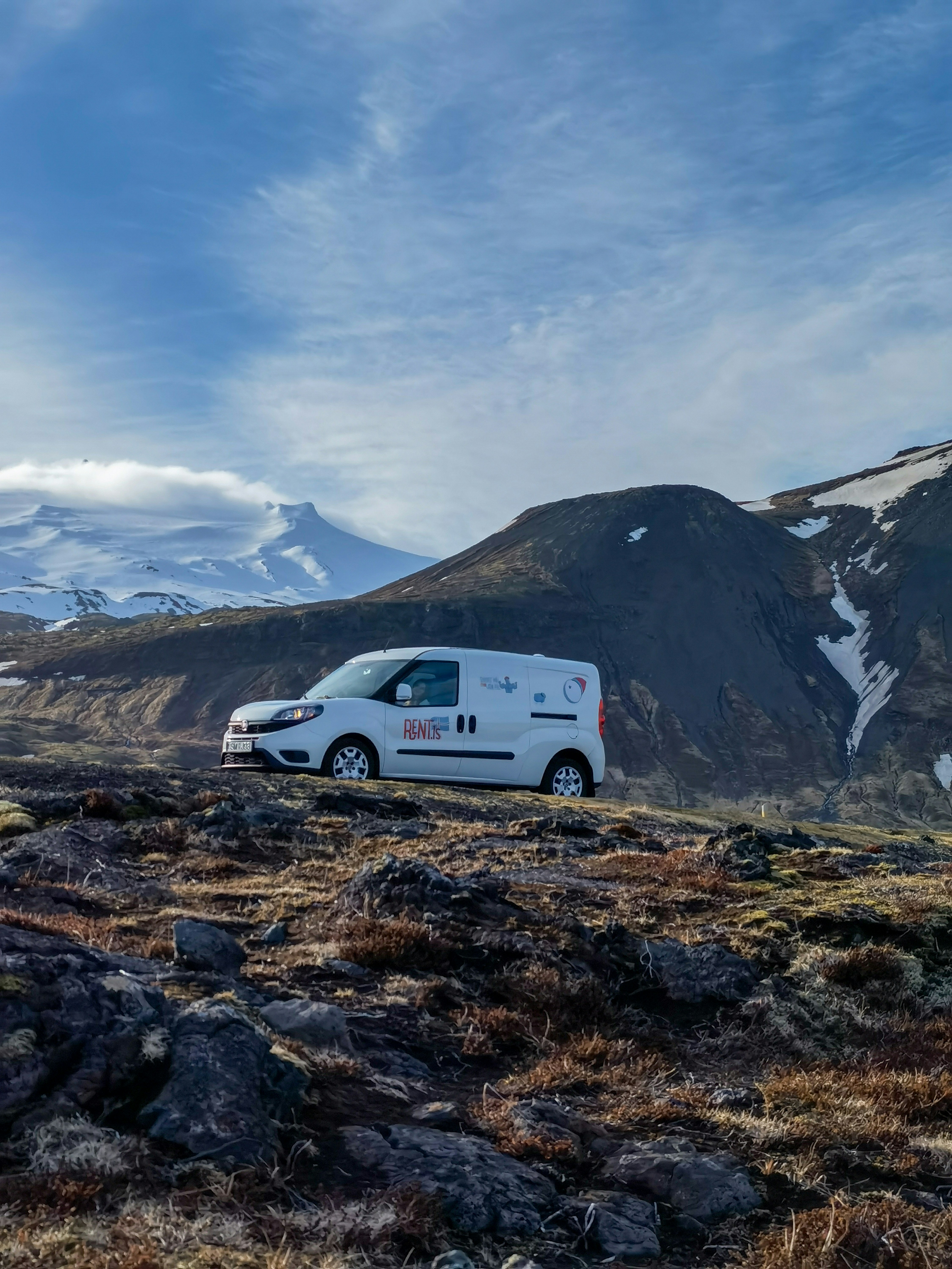 White utility van sits on rocky terrain with distant snow-capped peaks under a clear blue sky.