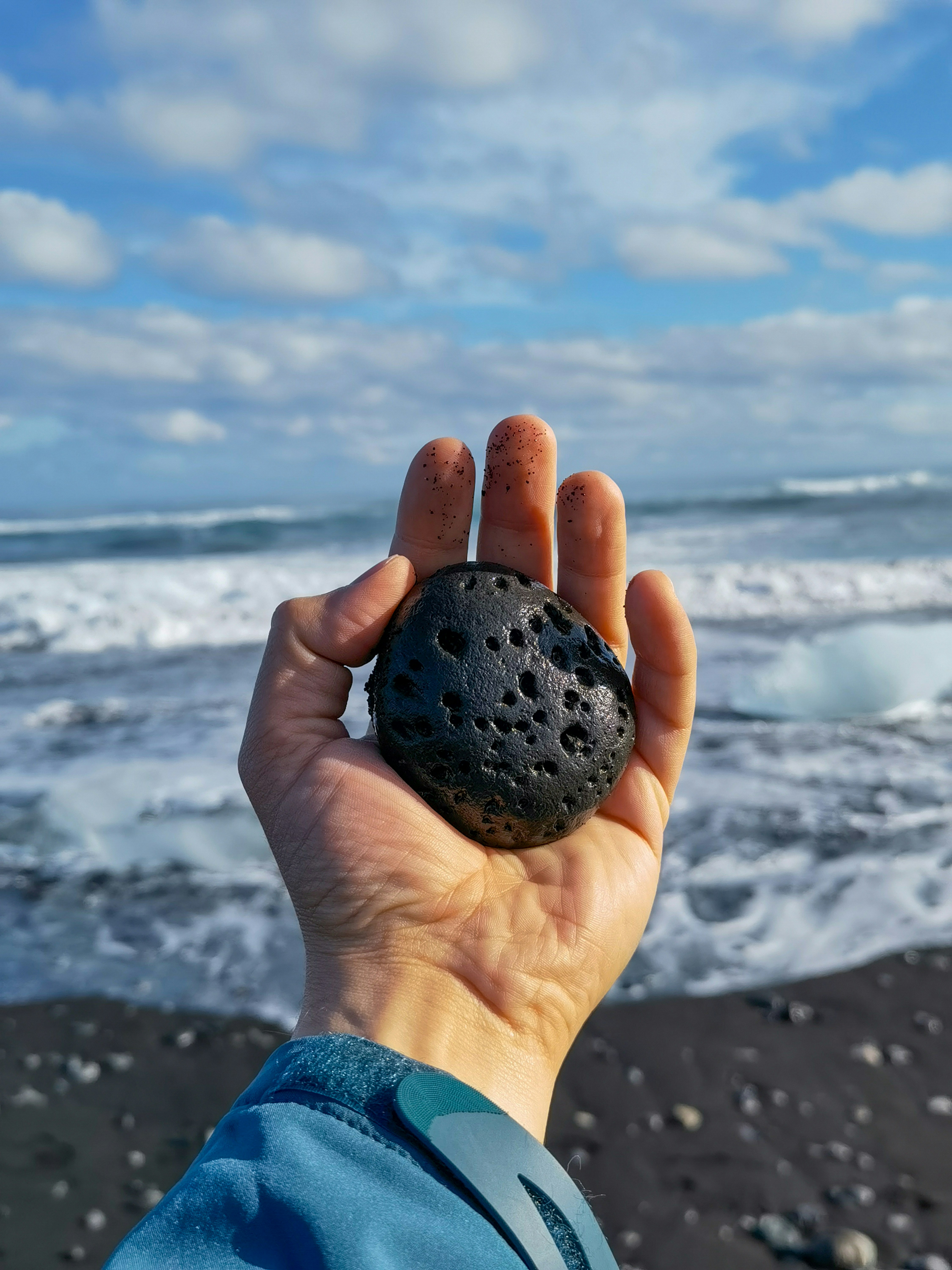Close-up of a hand gripping a porous black pumice rock, with foamy waves and blue sky in the background.