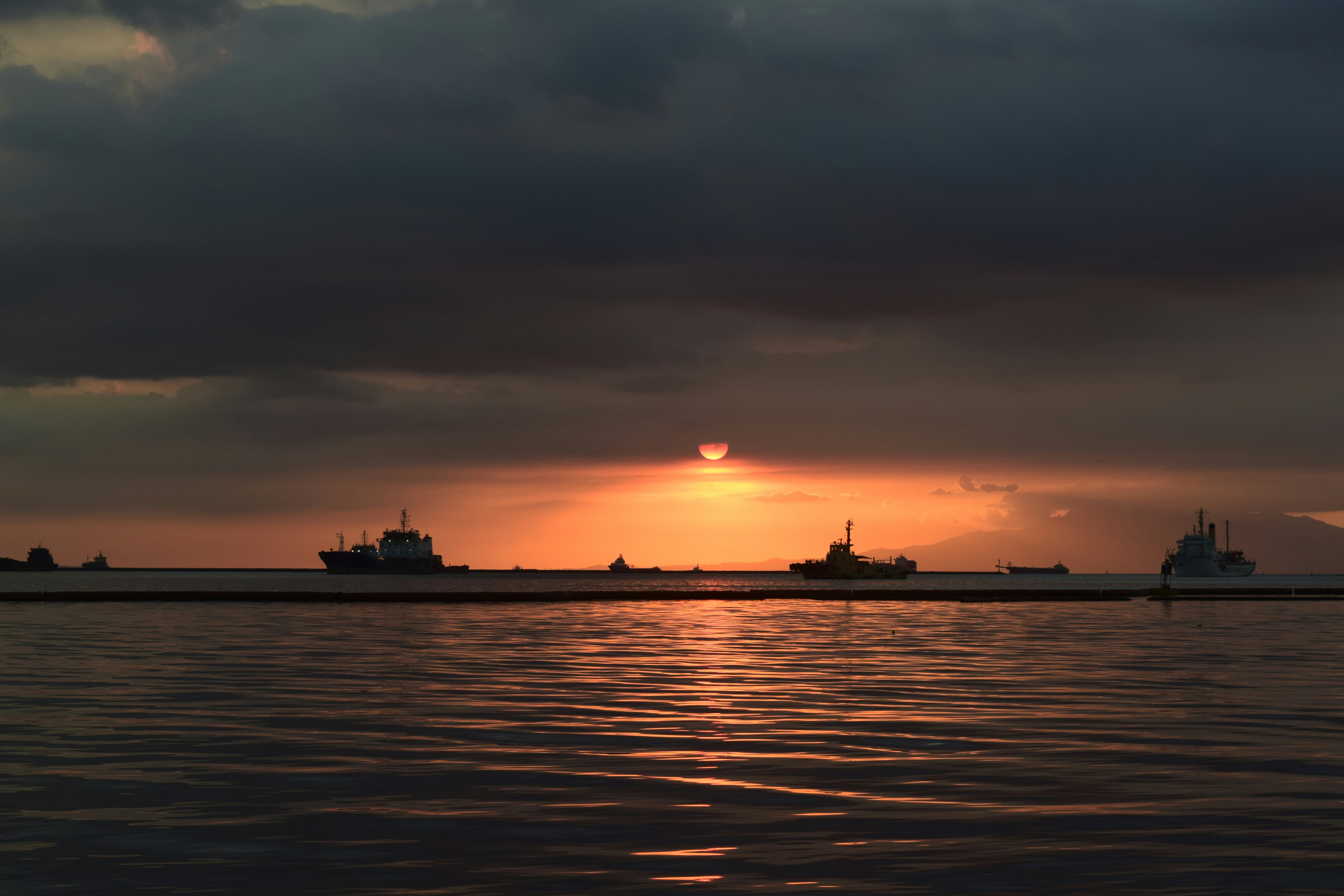 Sunset over calm waters with silhouetted ships on the horizon beneath a dramatic sky.