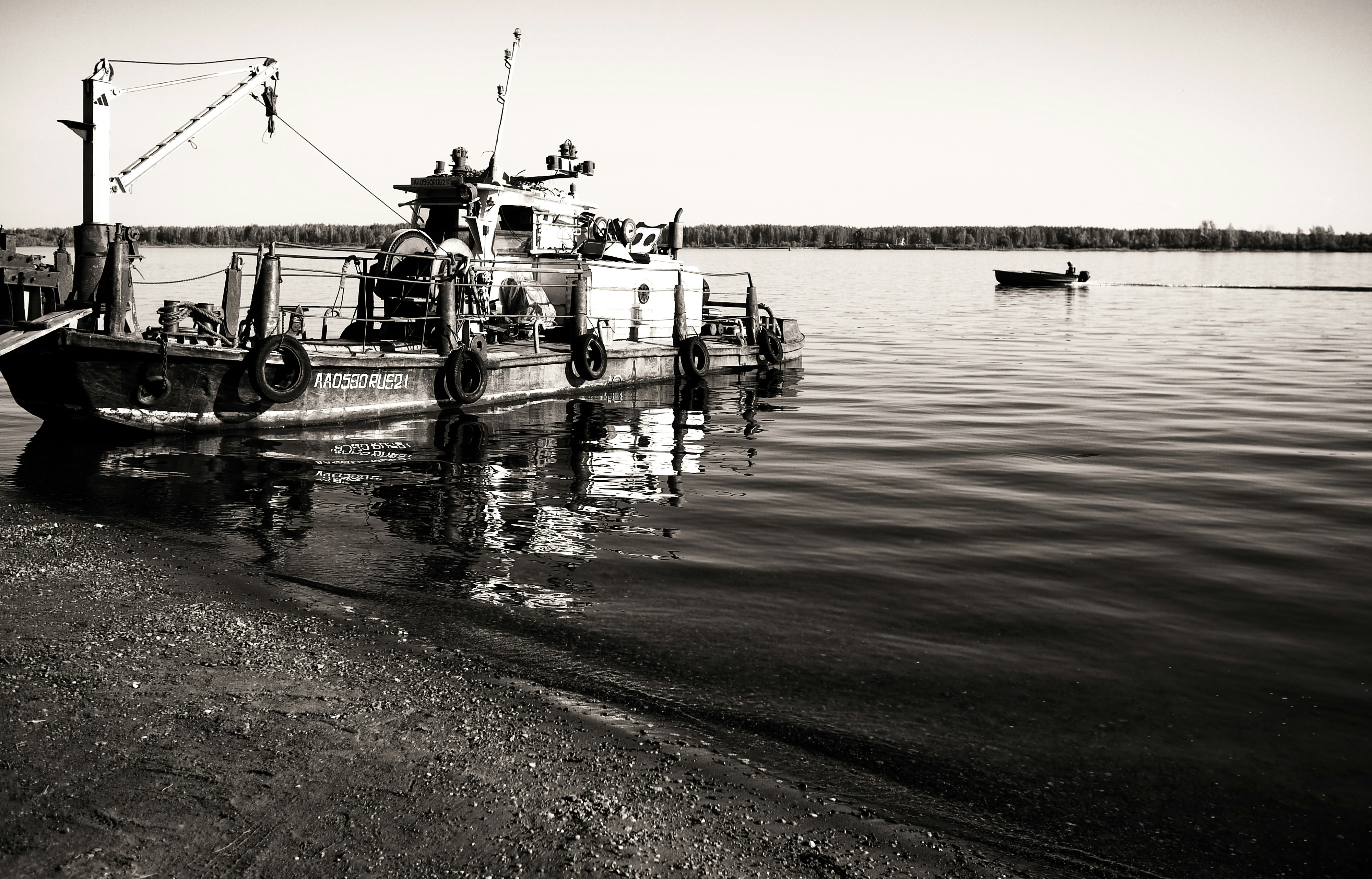 Black-and-white photograph of a weathered tugboat moored along a pebbled shoreline, with calm water stretching toward the horizon. A distant boat sits under a pale sky.