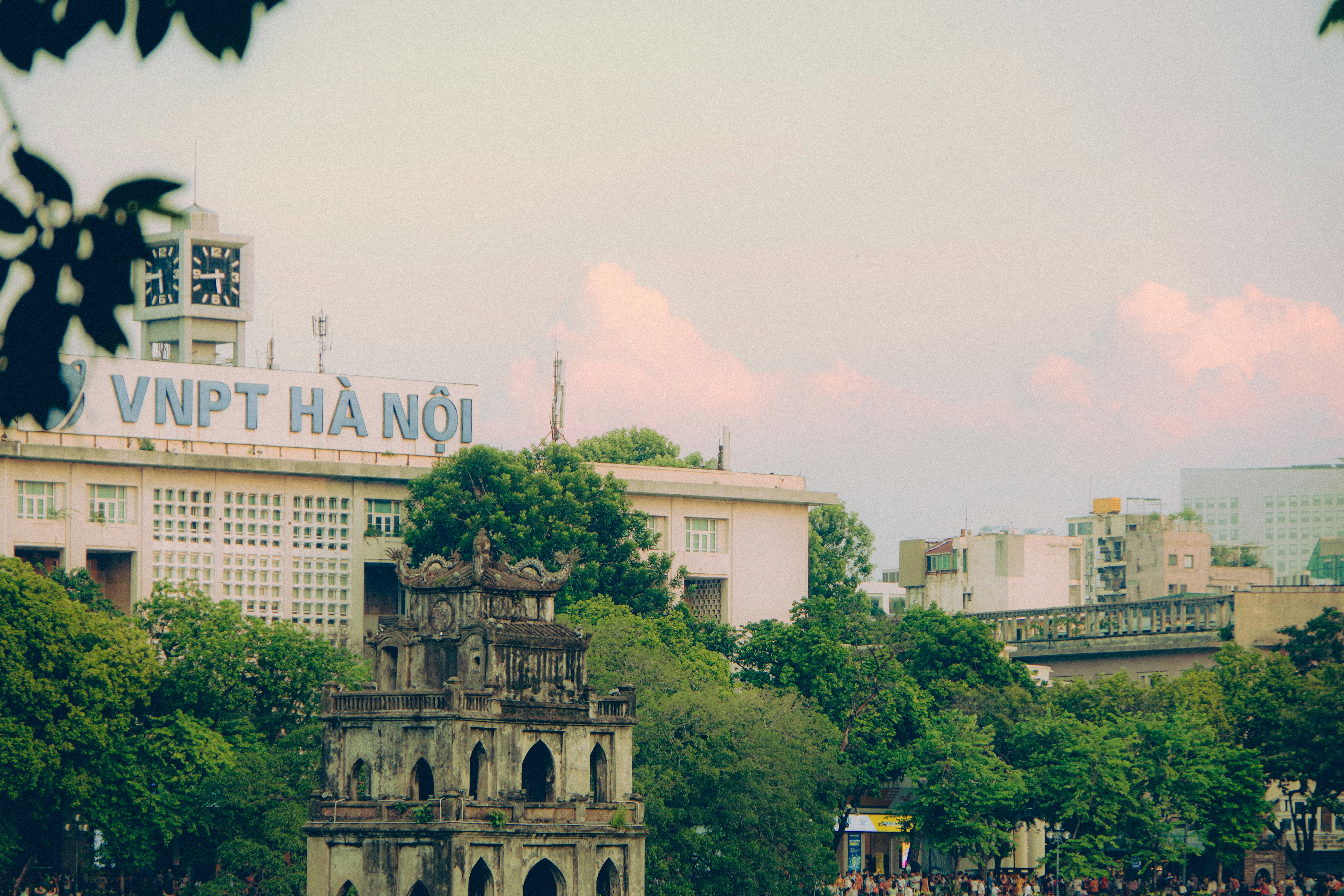 The iconic Turtle Tower rises amidst lush greenery, with the VNPT Hà Nội building in the background, capturing a blend of history and modernity in the bustling city.