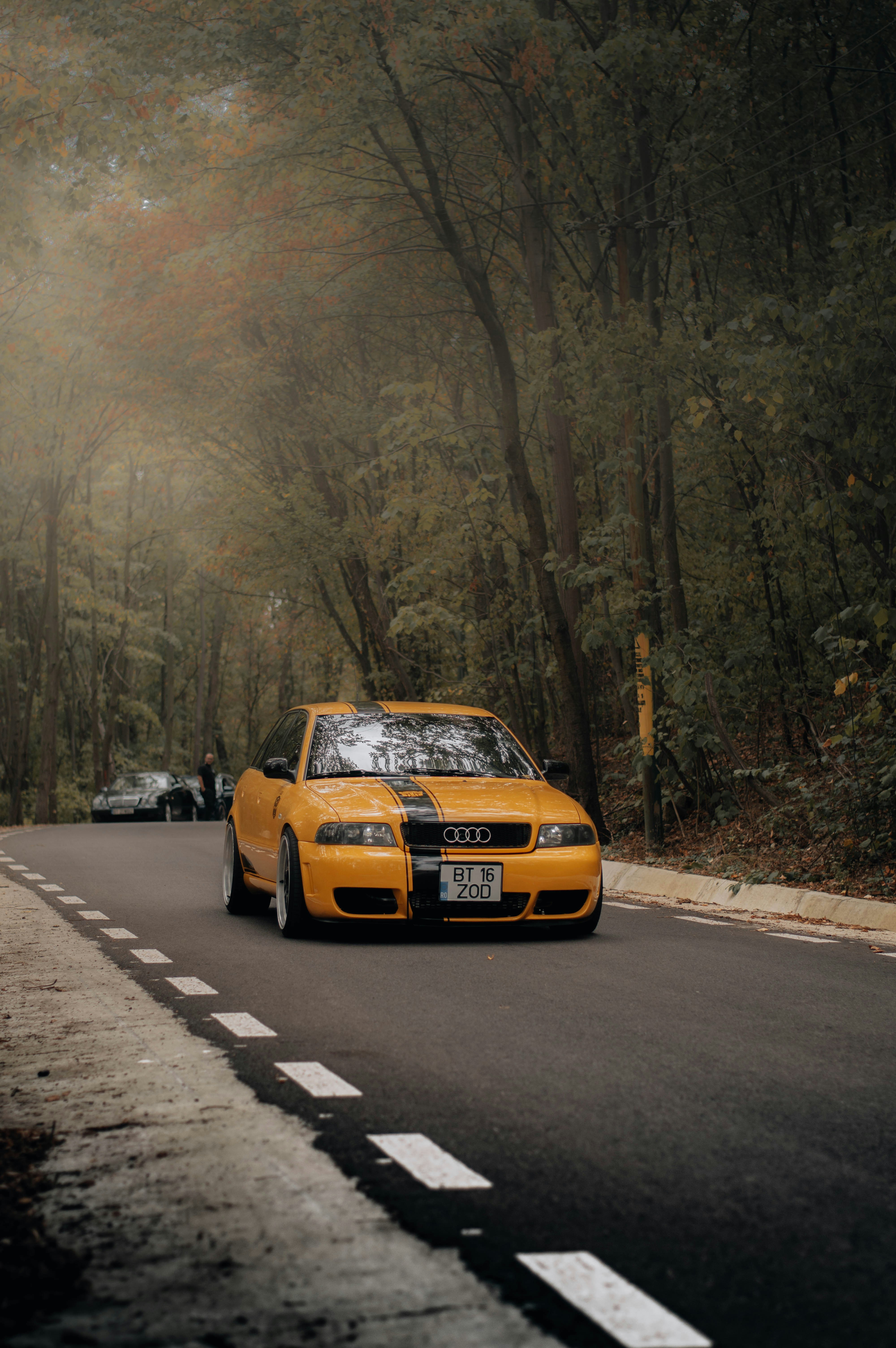 A yellow car driving down a road next to a forest photo – Free Car ...