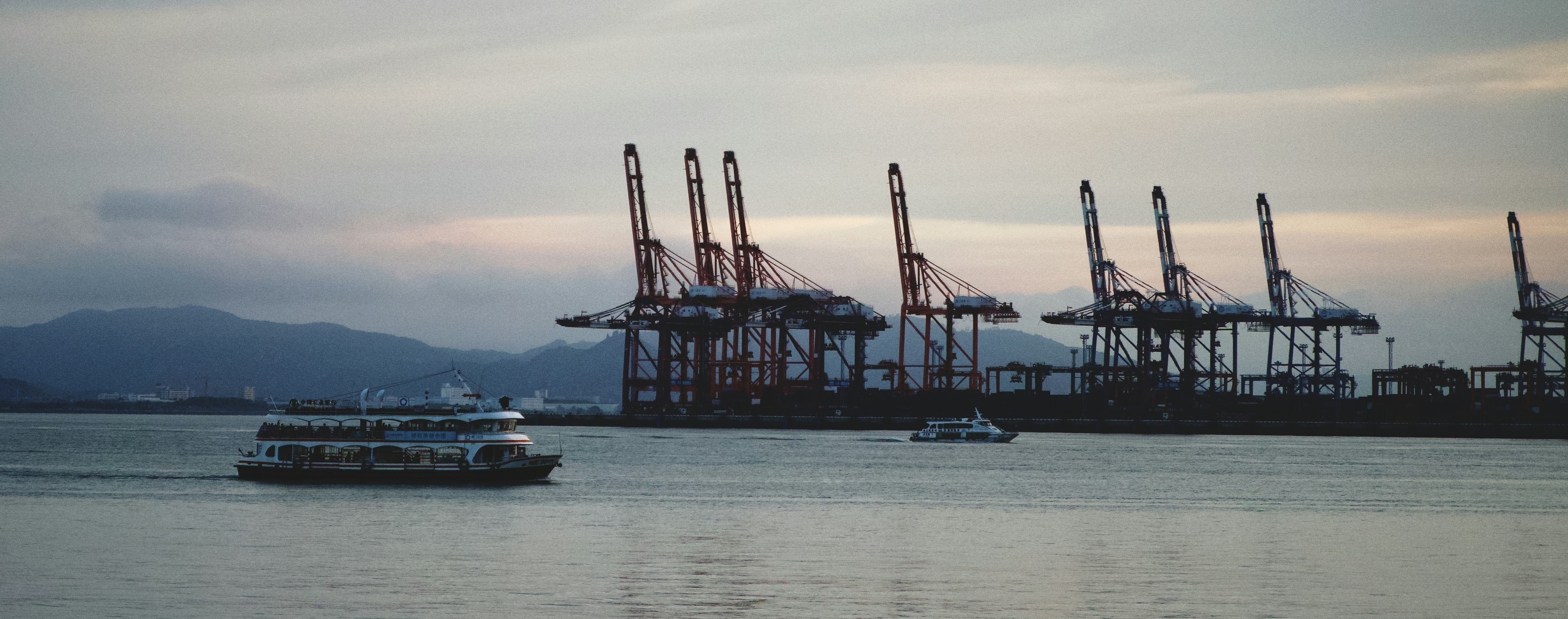 Boat gliding toward a pier with towering cranes silhouetted against a pastel sky.