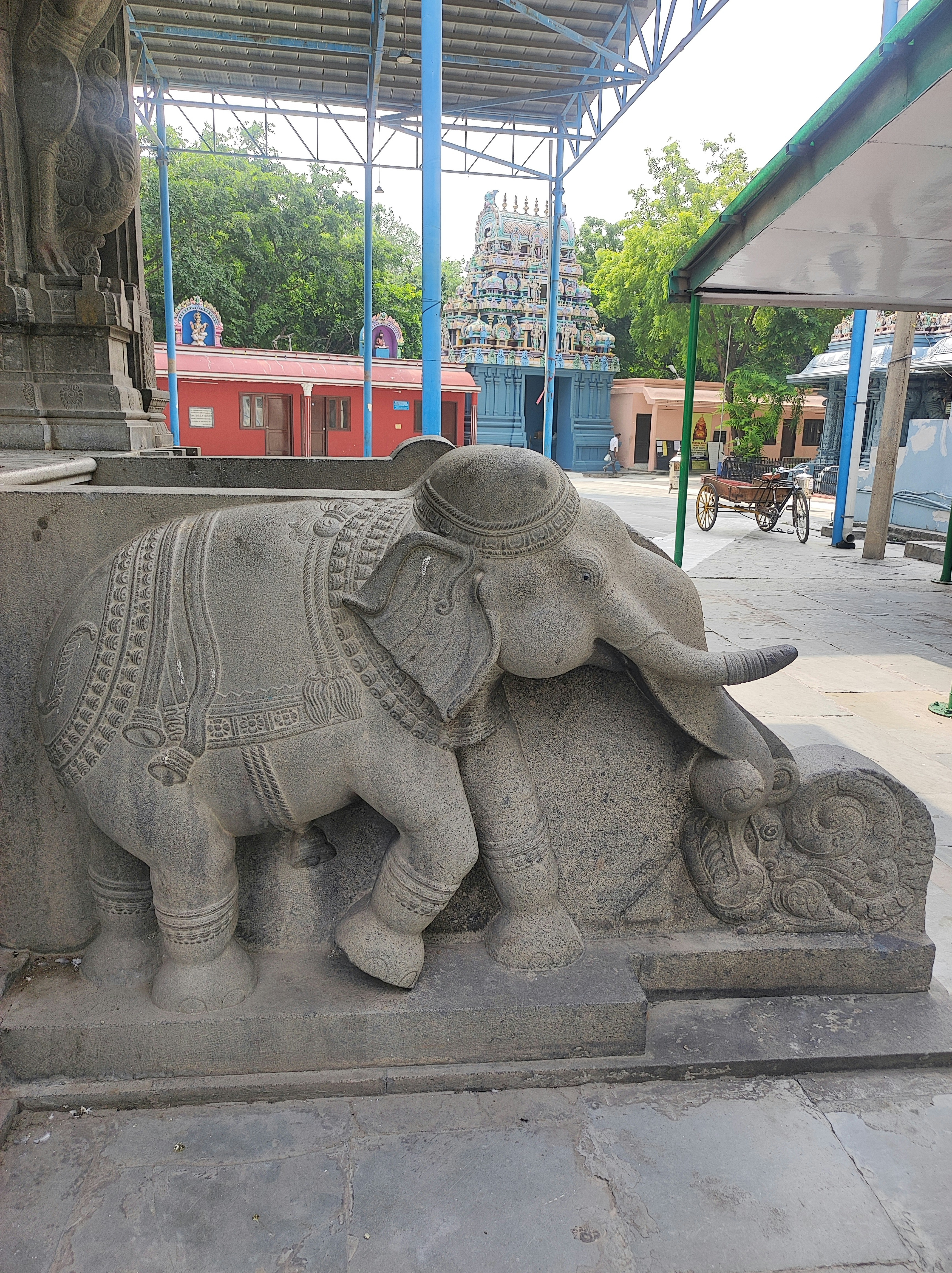 Ornate stone elephant sculpture guards a temple courtyard beneath a metal canopy, with distant temple structures and a bicycle visible in the background.