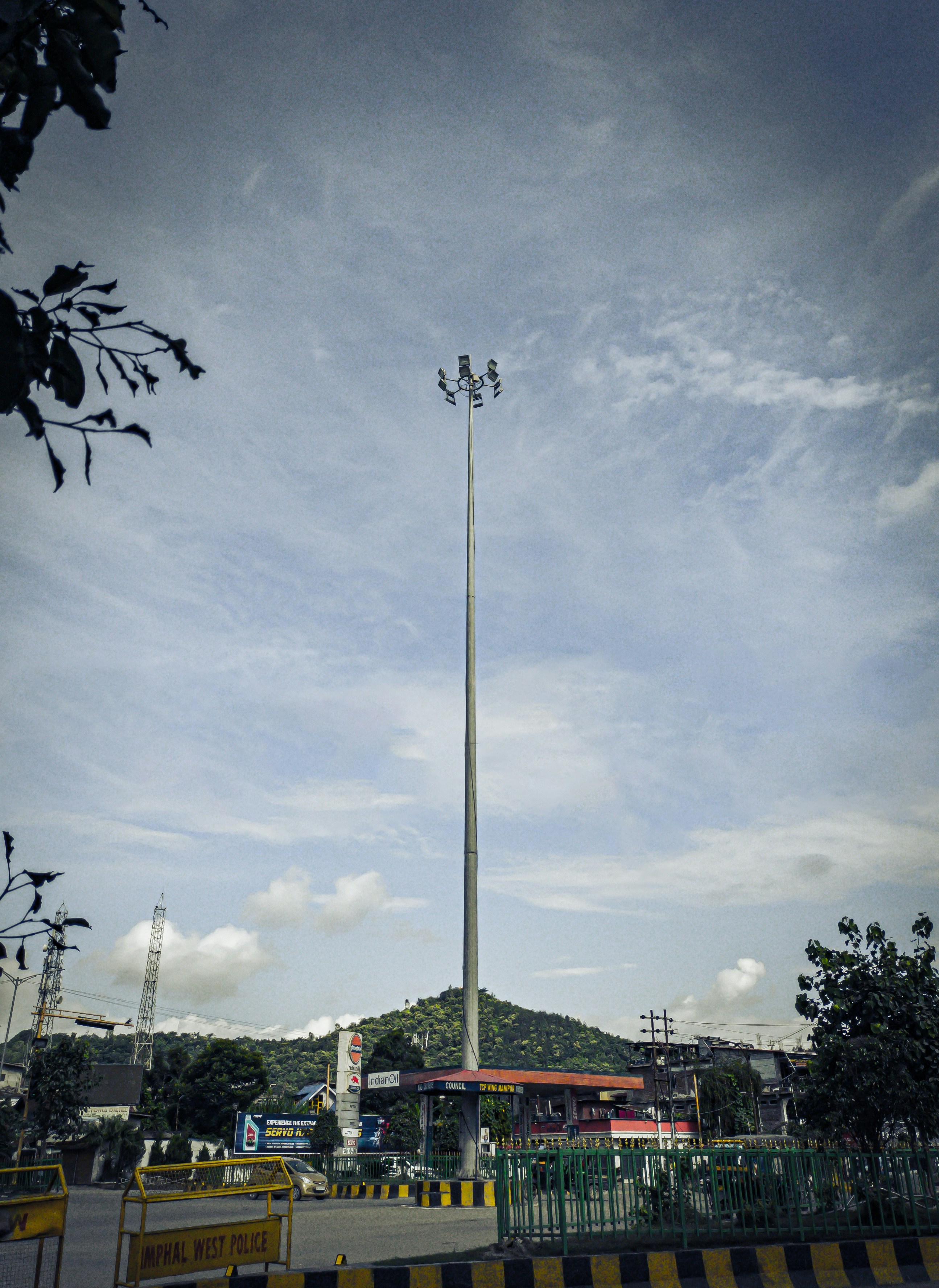 Tall streetlight mast rises over a municipal roundabout beneath a partly cloudy sky. The foreground features road barriers and a distant hill, with the pole forming a stark vertical centerpiece.