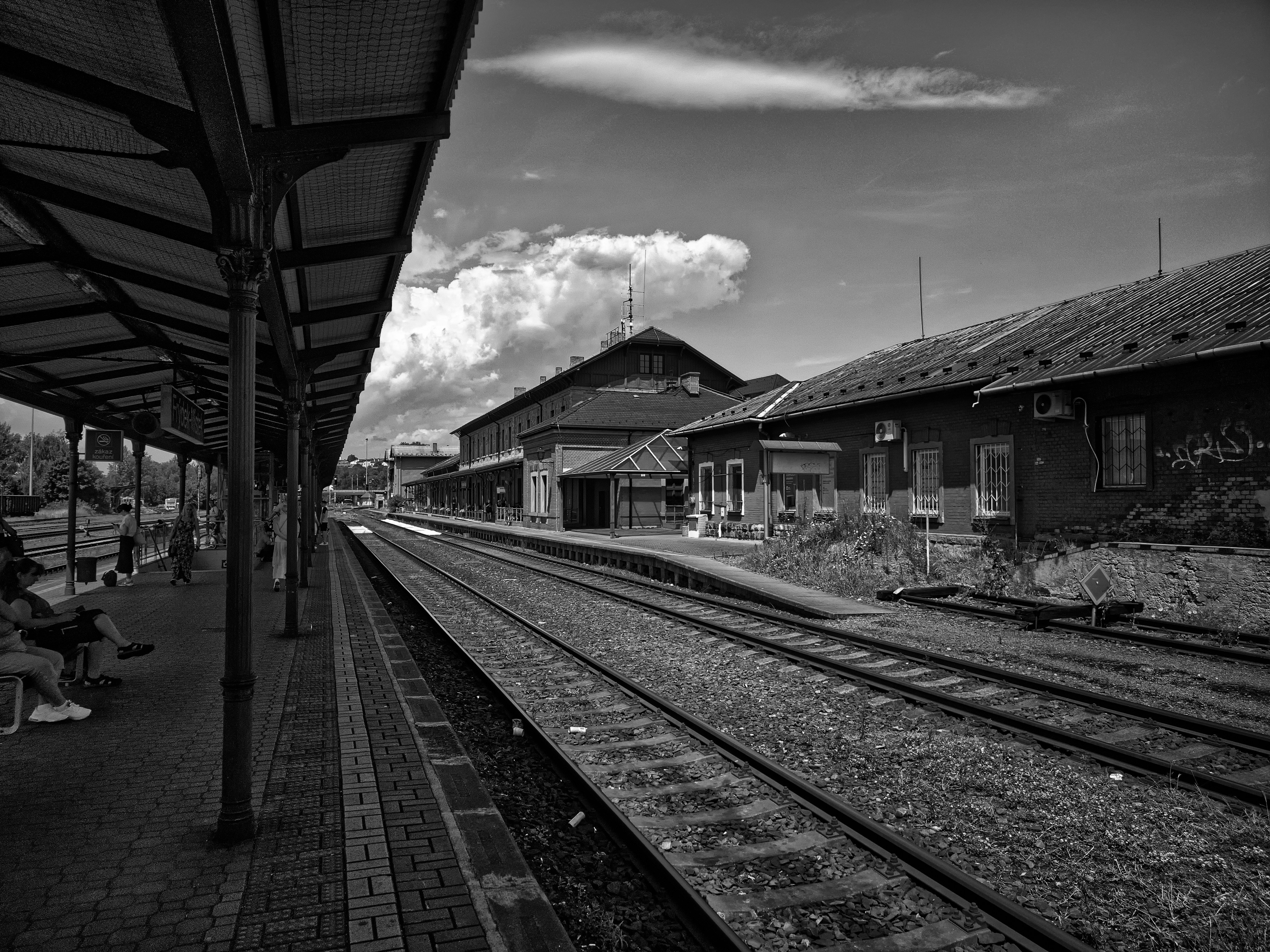 Black-and-white photograph of a train platform with parallel tracks converging toward a distant station. A weathered building lines the right edge while a few passengers sit along the left.
