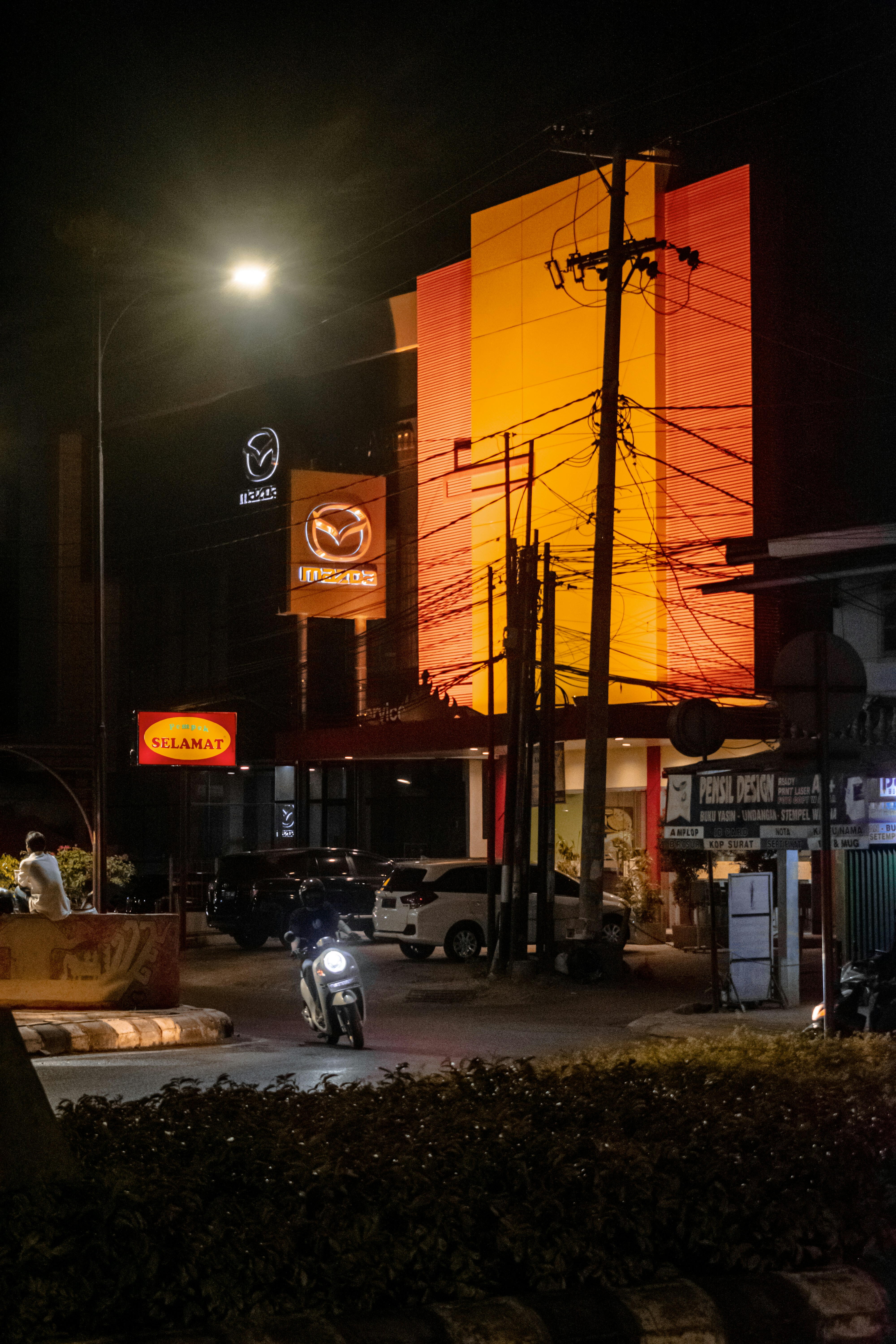Orange Monolith at Night: Mazda Sign, Scooter, and Neon Poles