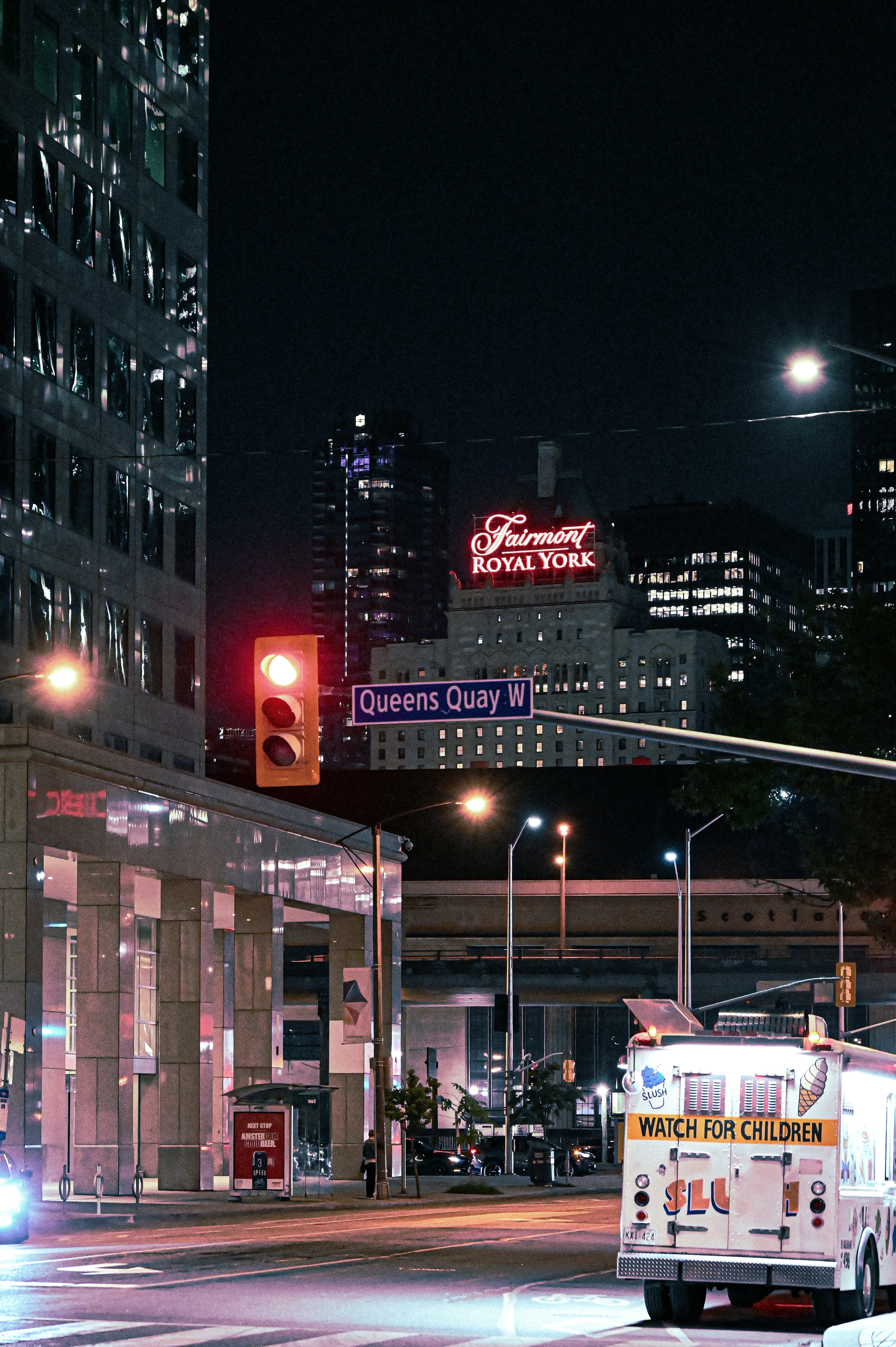 Illuminated street scene featuring the Fairmont Royal York sign and traffic lights, with a vintage streetcar in the foreground. Nighttime urban atmosphere captured with vibrant colors.
