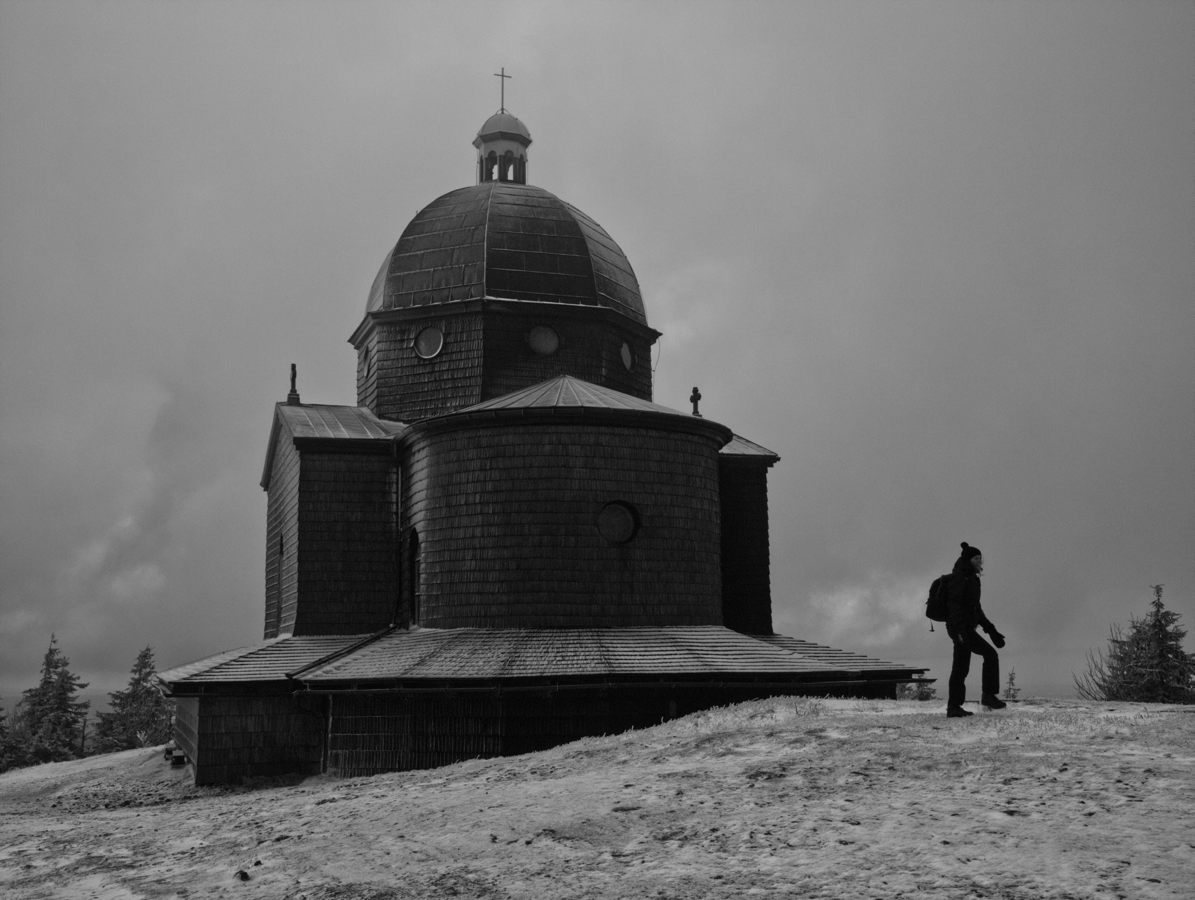 Black and white scene of a solitary figure standing near a domed chapel under a cloudy sky.