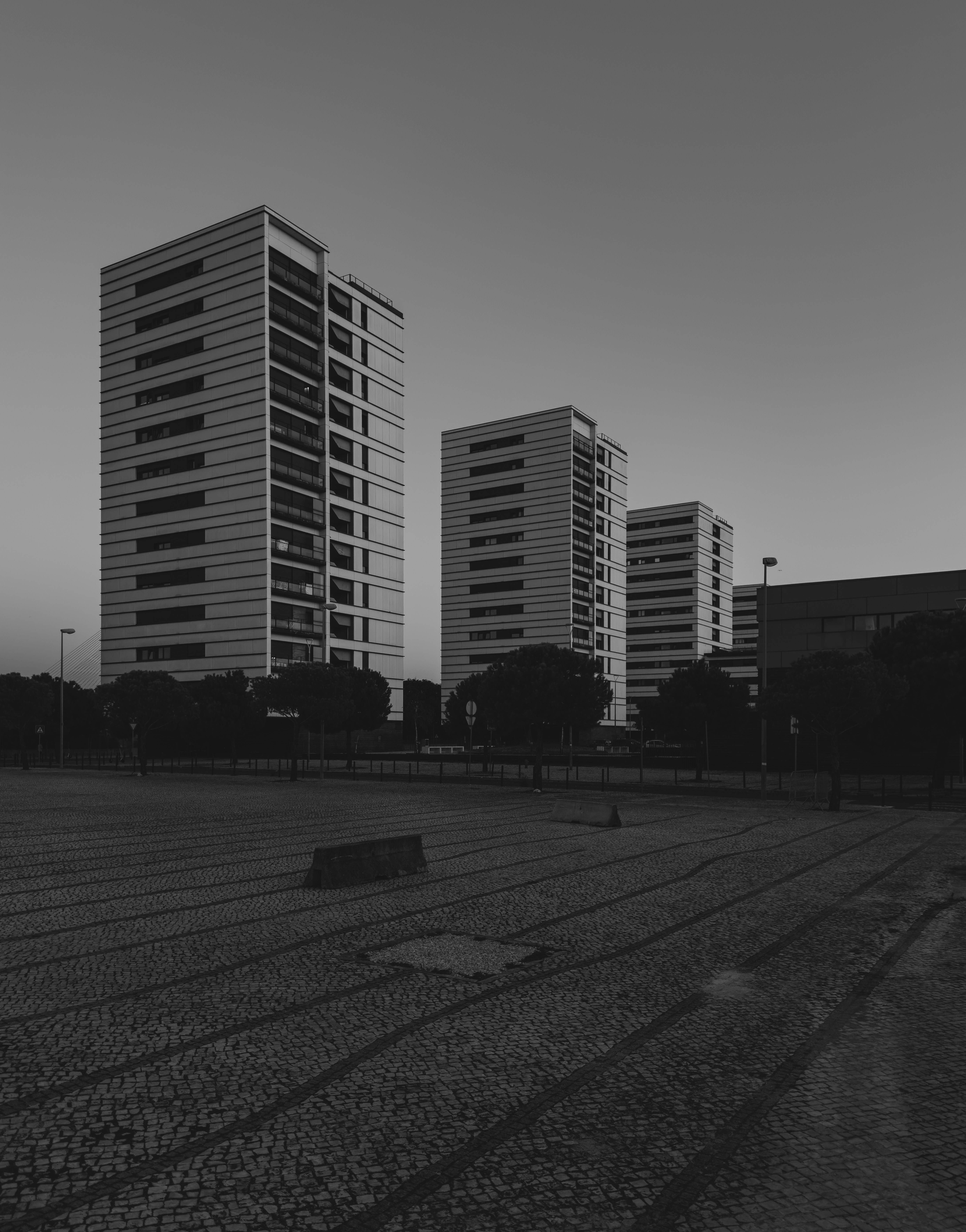 Grayscale photograph of three modern high-rise blocks rising above a cobblestone plaza beneath a clear sky.