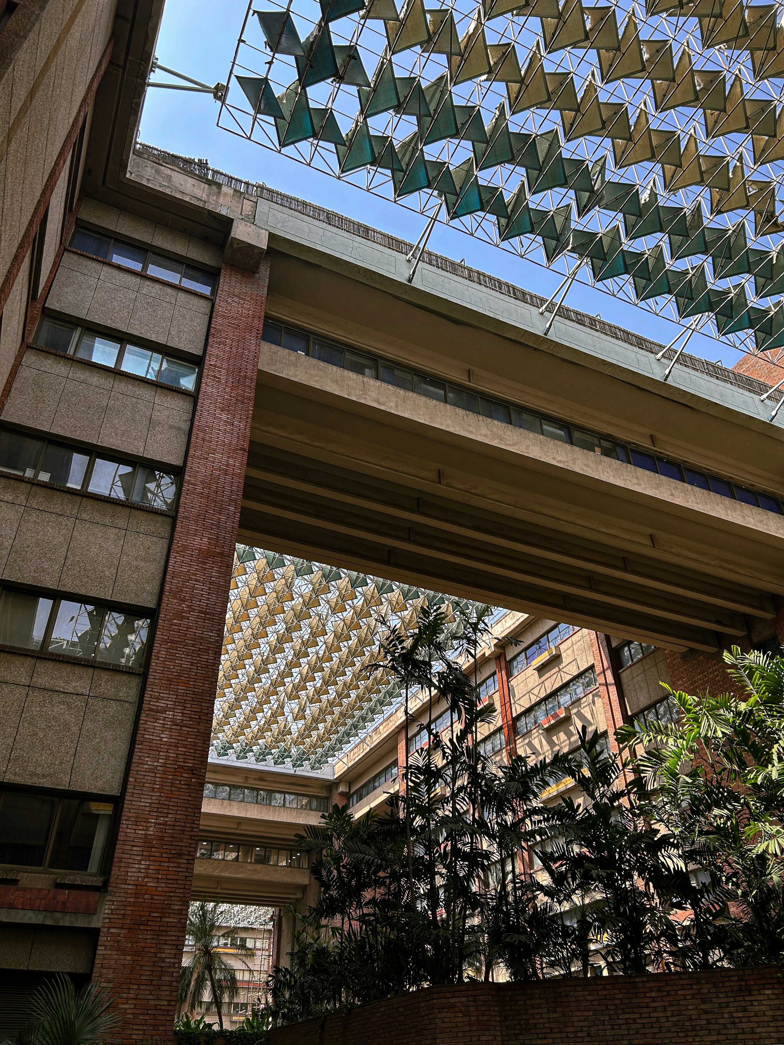 Photograph of an urban courtyard featuring a diamond-pattern glass canopy over brick and concrete walkways, with lush greenery below.
