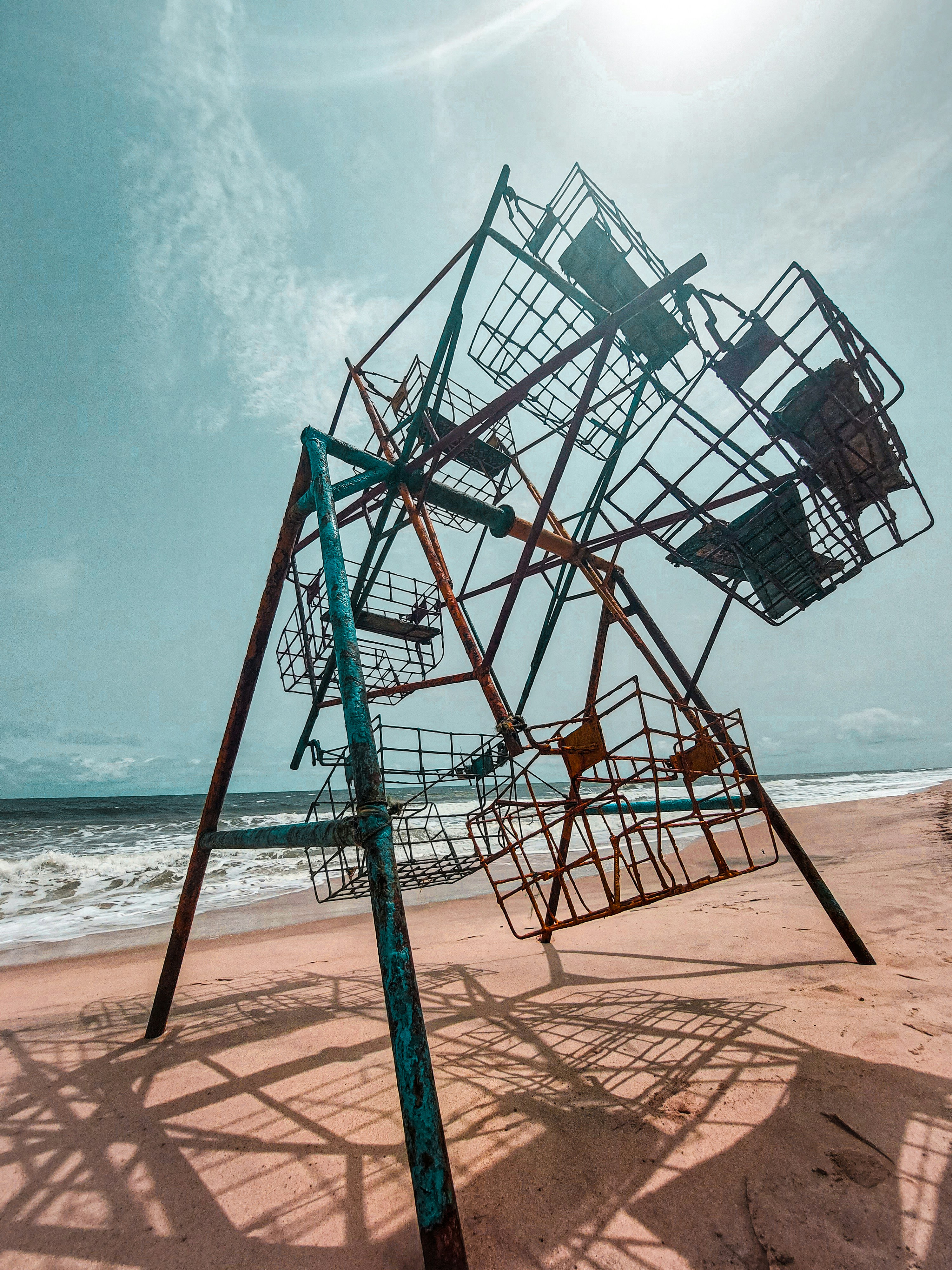 Geometric rusted-metal sculpture leans on the sandy shore, its angular grids casting intricate shadows as the ocean and bright sky unfold in the background.