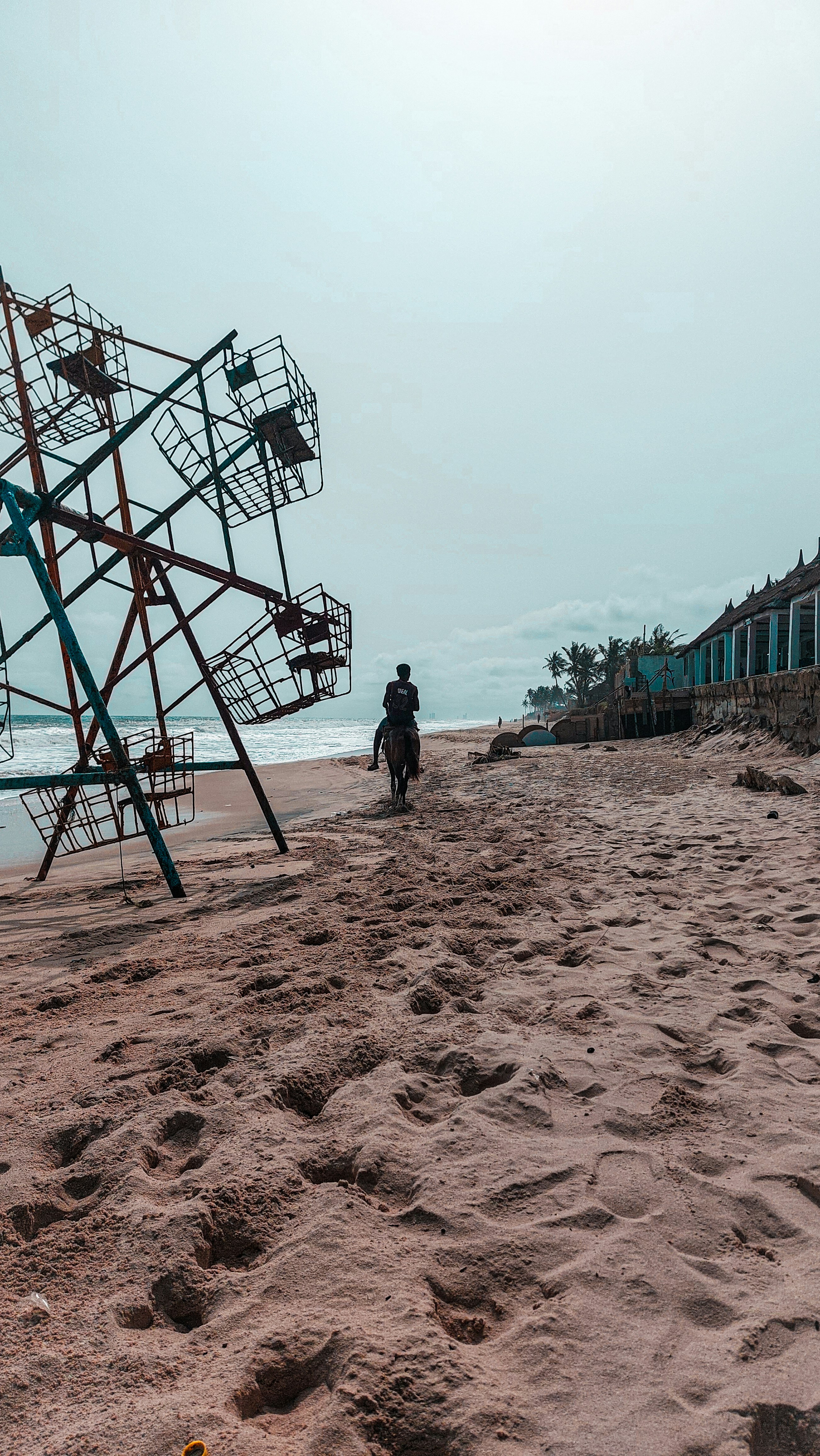 Photograph of a lone figure walking beside a bicycle along a sandy beach, with a skeletal ferris wheel frame on the left and a calm ocean on the horizon.
