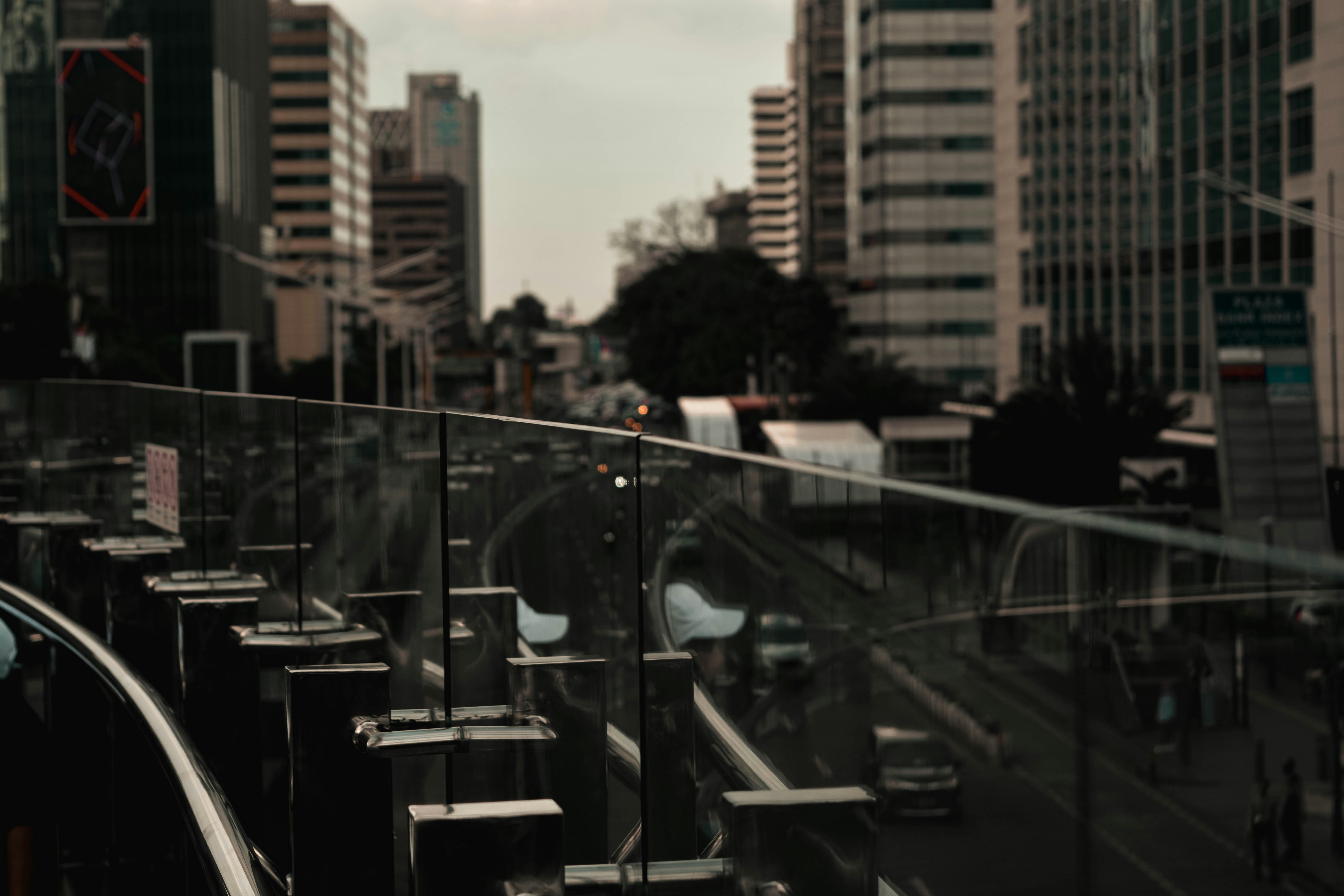 Cityscape featuring a busy street viewed through a glass railing, with towering buildings in the background. The scene captures the essence of urban life.