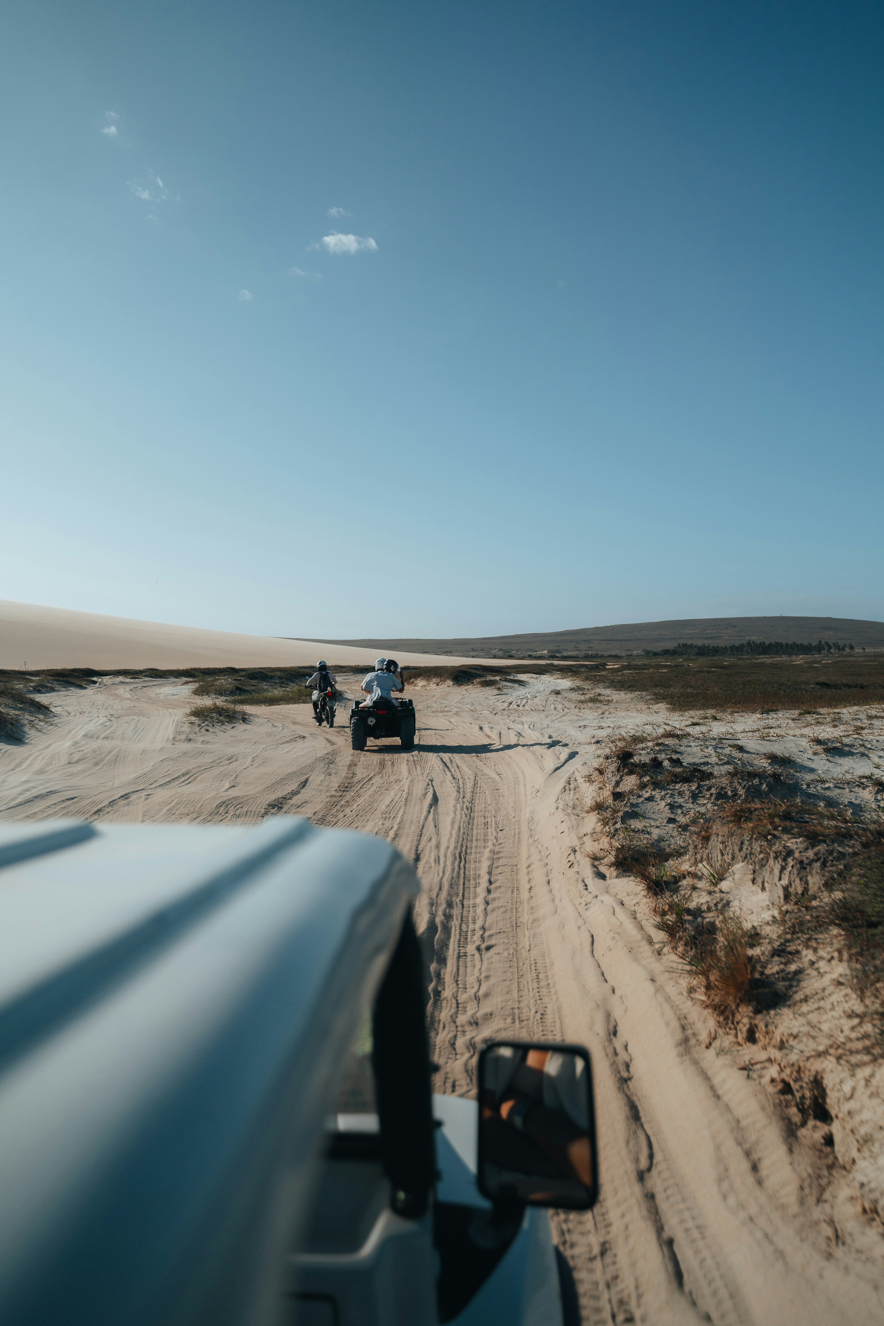 Two off-road vehicles navigating a sandy path, with a vast landscape and distant hills under a clear blue sky.